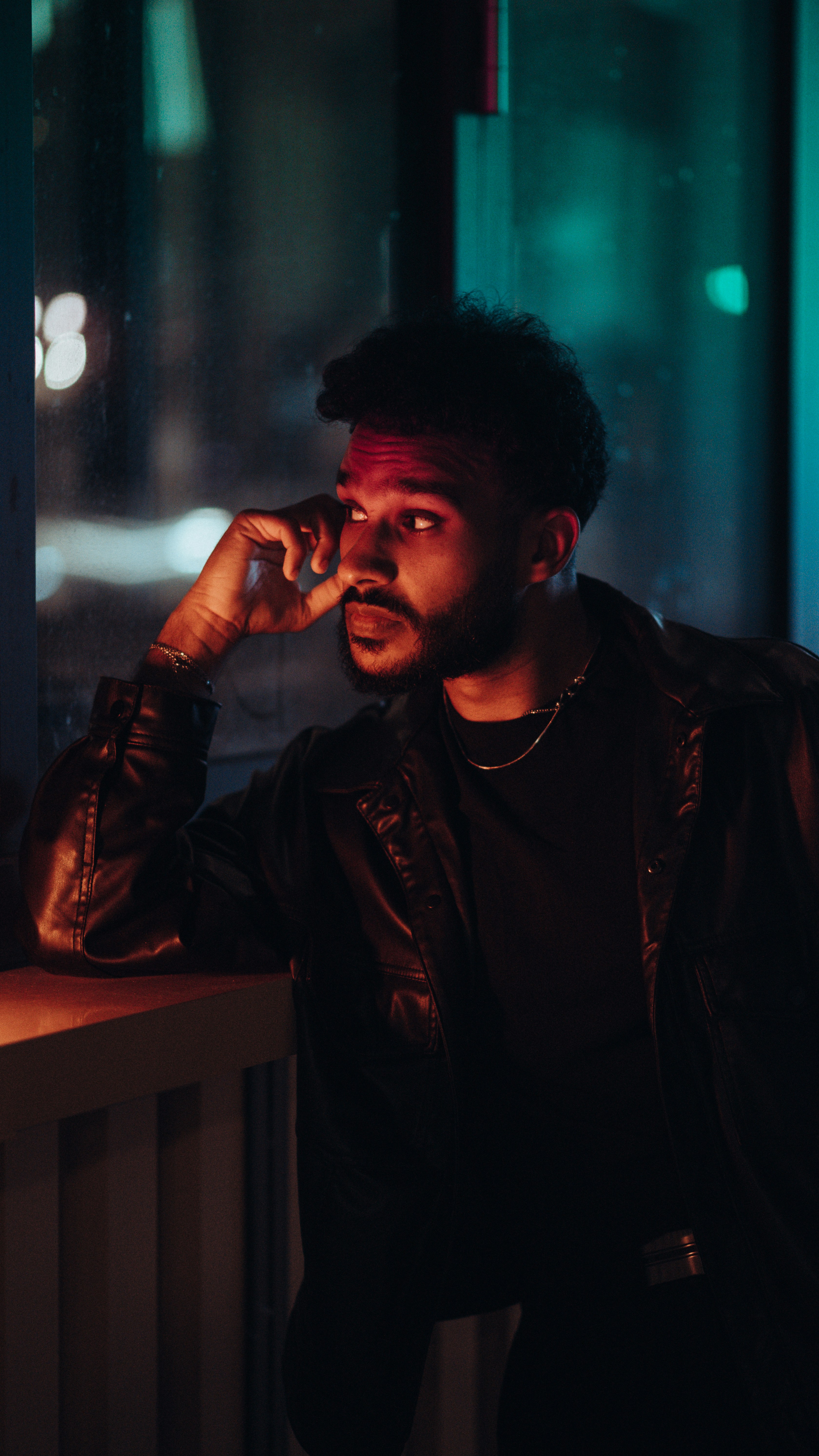 A contemplative urban portrait shot through a window at night. The subject looks away in thought, bathed in neon city lights, giving the image a mysterious, late-night atmosphere perfect for album art or storytelling.