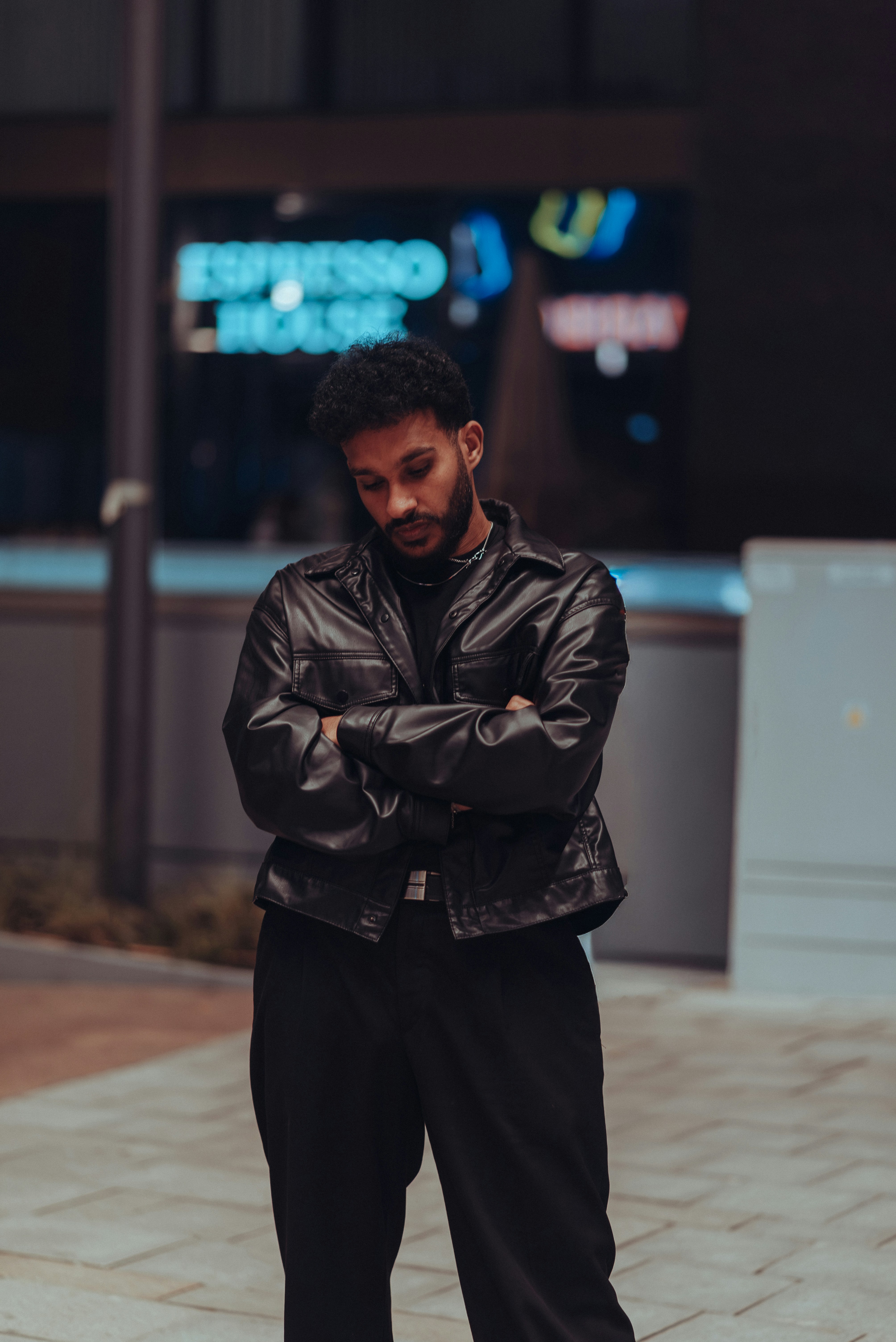 A fashionable man standing on a city street at night with arms crossed. He is wearing a black leather jacket and looking down. The background features blurred blue neon city lights and bokeh, creating a moody urban atmosphere