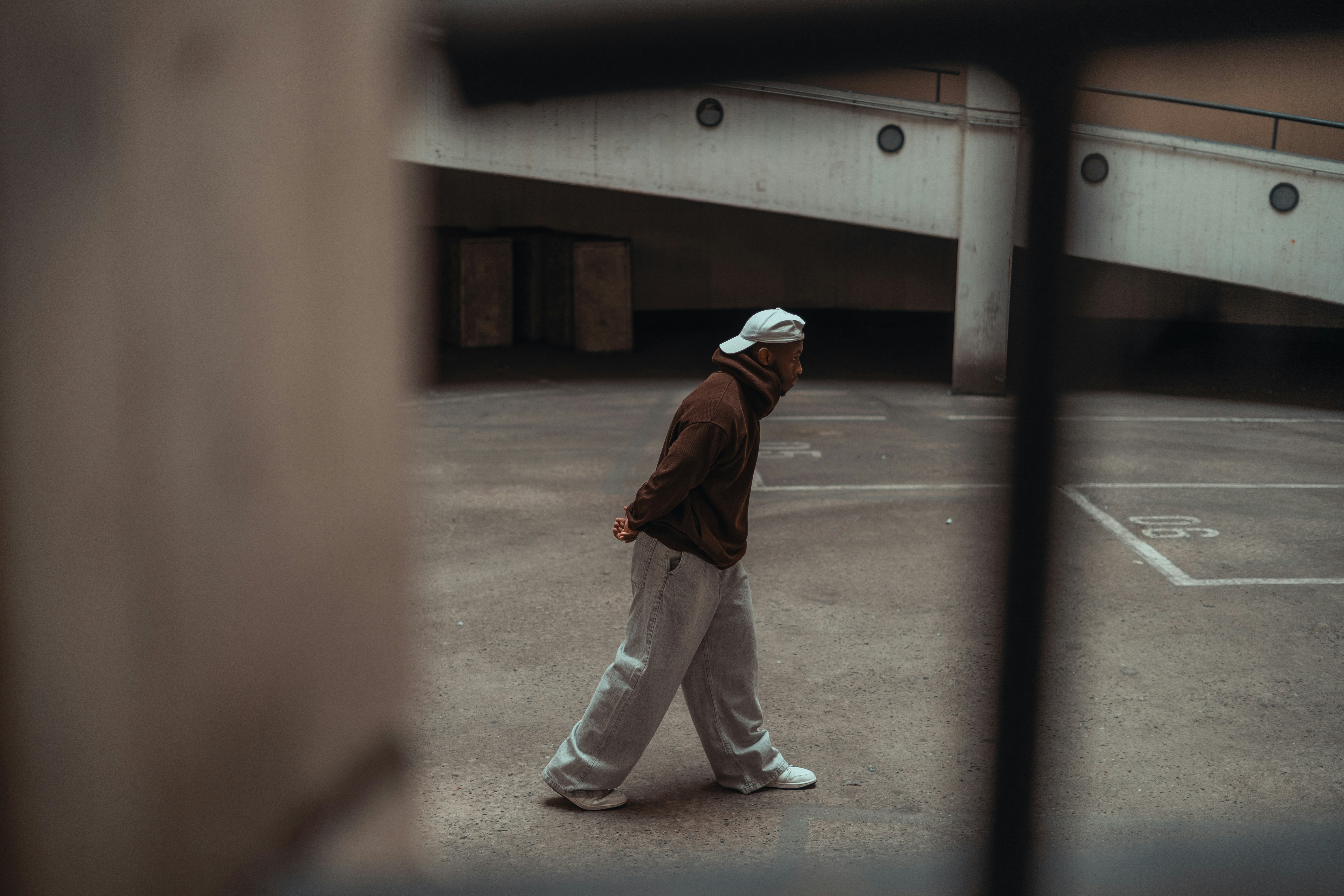 Candid side profile of a man walking through a concrete parking structure, viewed through foreground bars or fencing. The natural framing and muted tones create a sense of urban exploration and mystery.
