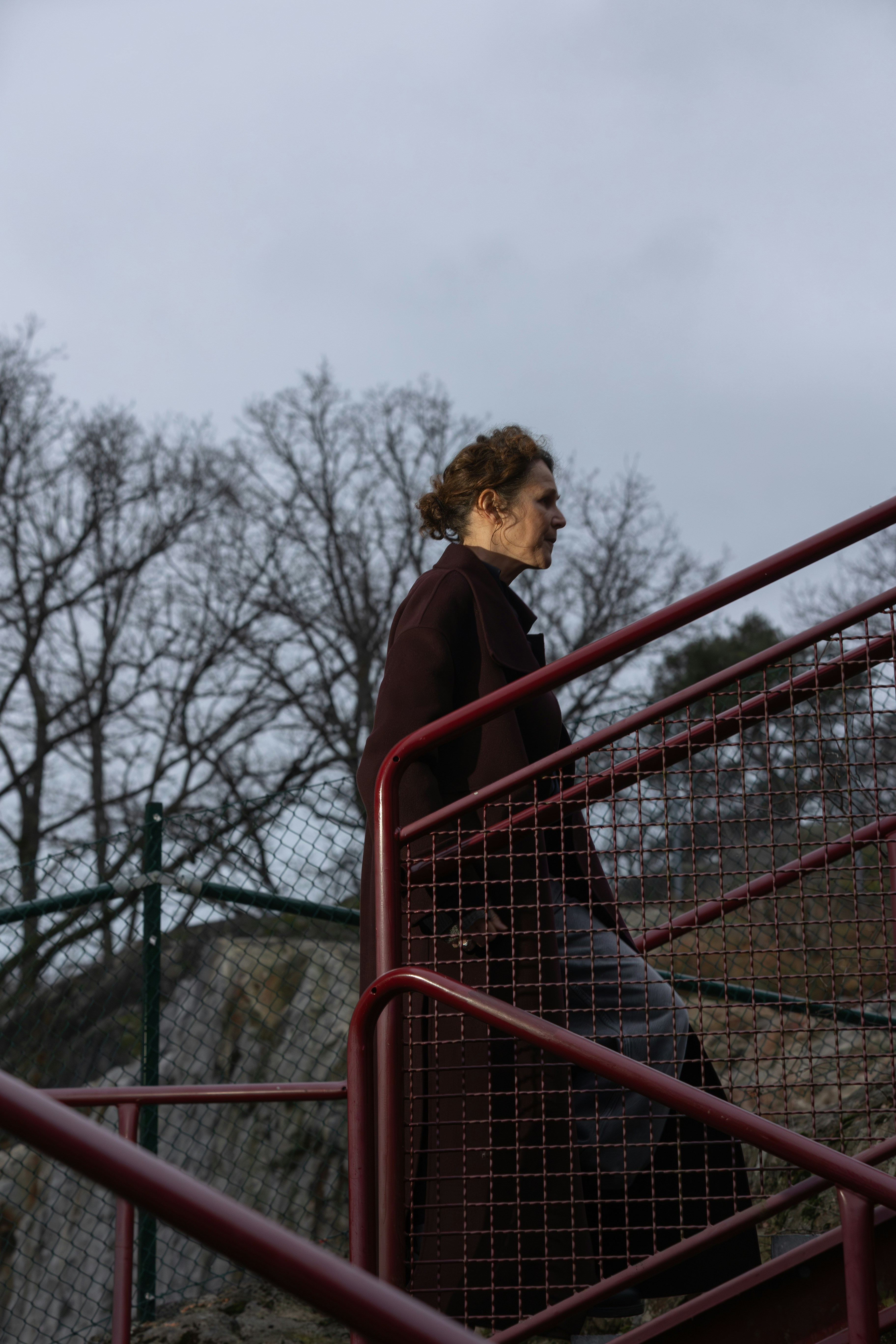 A woman wearing a maroon colored overcoat climbs a metal outdoor staircase in a December gloomy Stockholm.