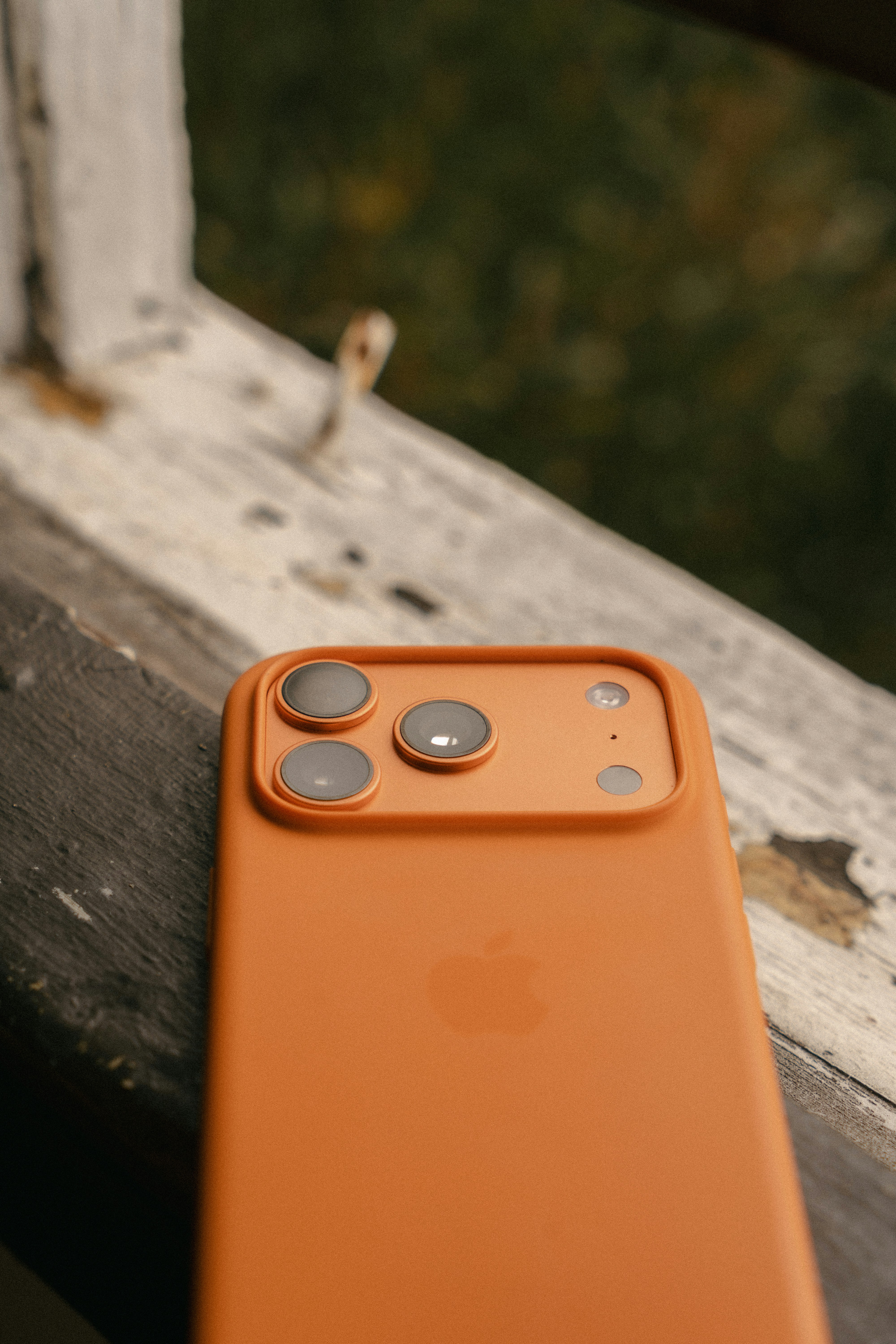 Close-up detail of a smartphone camera wrapped in a soft matte orange case, resting on a weathered windowsill. The contrast between modern technology and rustic texture creates a timeless editorial feel focused on design, photography, and everyday tools.