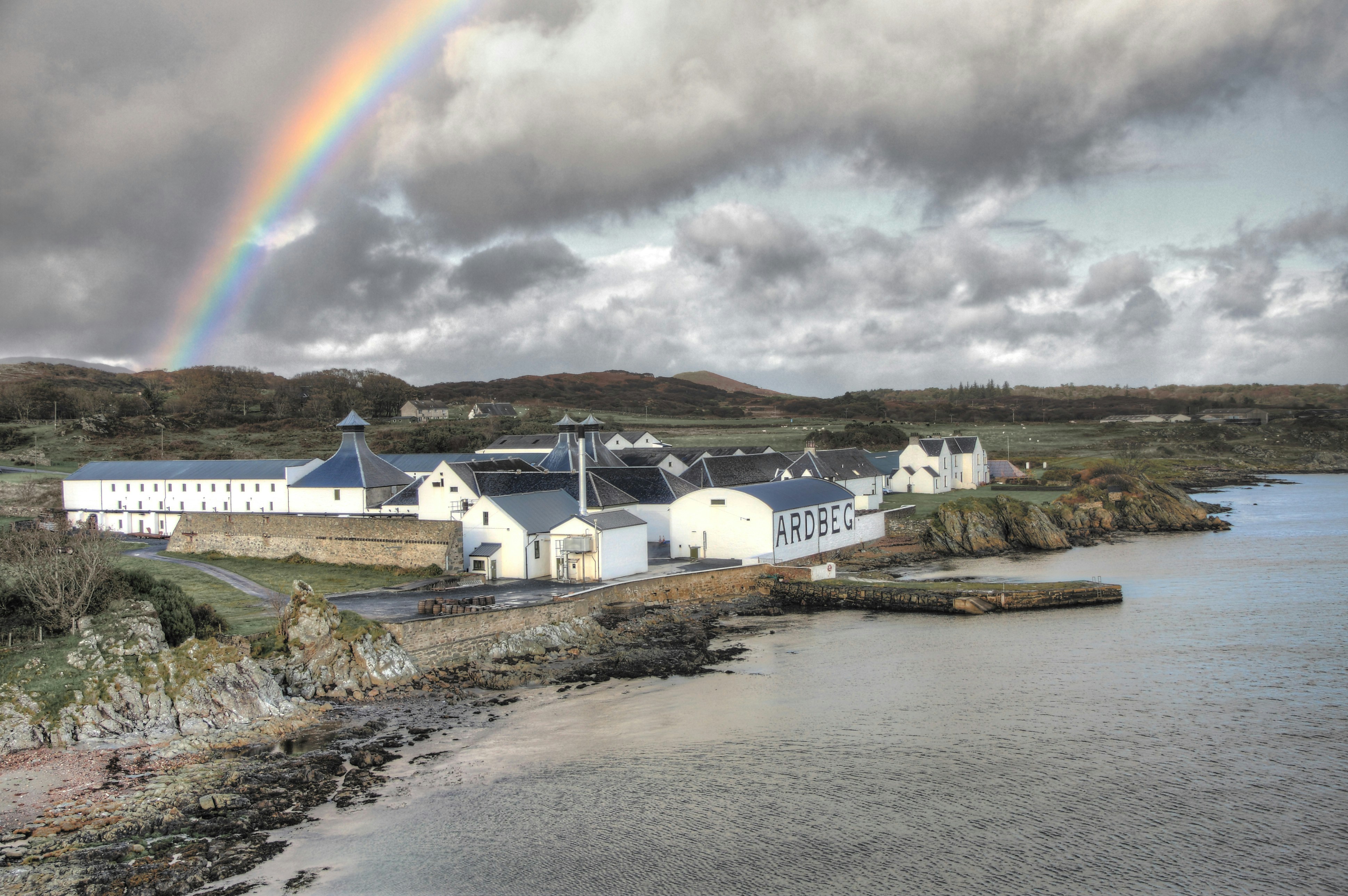 A rainbow arches over a coastal distillery under cloudy skies. photo ...