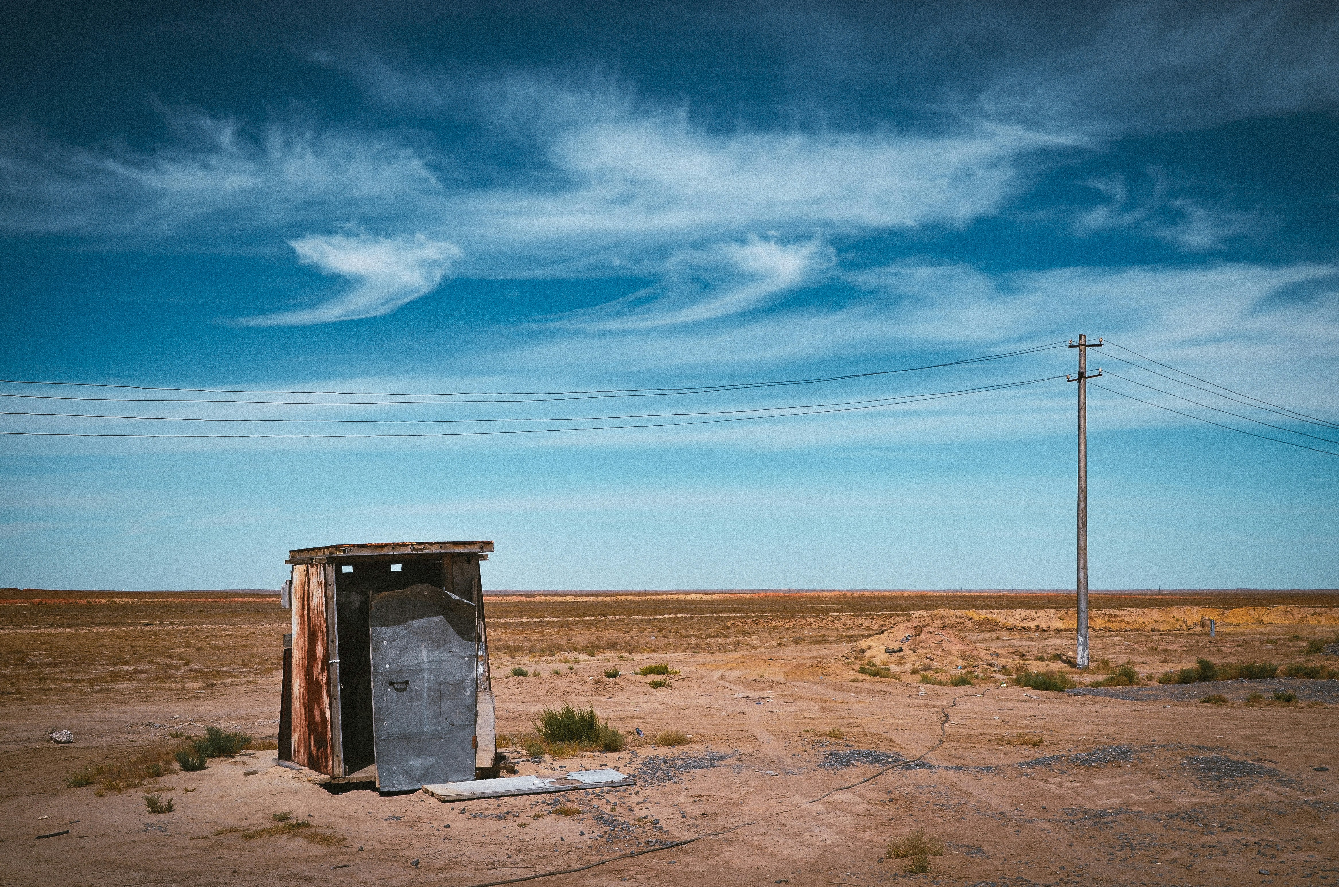 A small weathered shed stands alone in a barren landscape of cracked earth, framed by sparse vegetation and utility poles stretching into the distance.