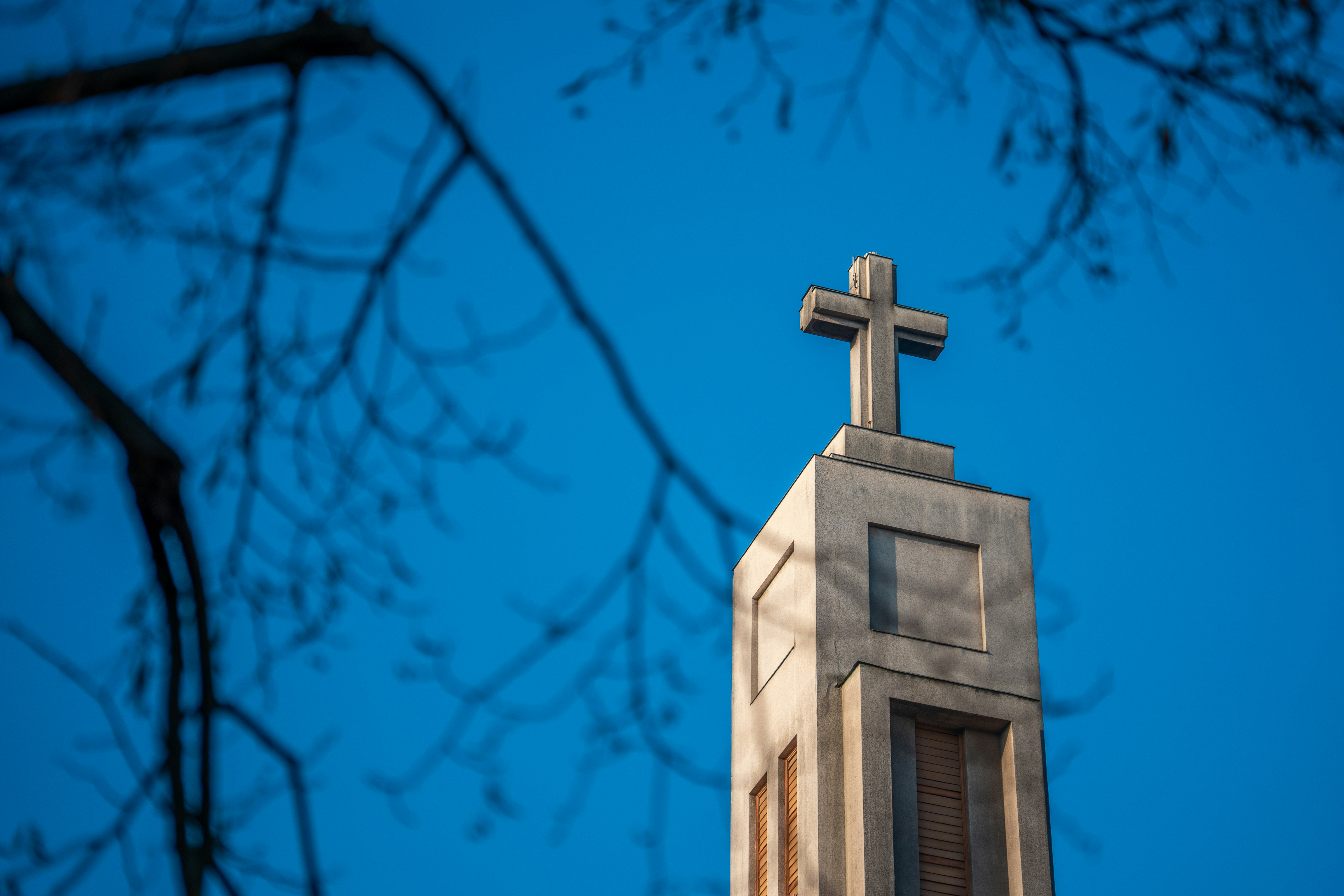 A church tower with a stone cross framed by bare tree branches against a deep blue sky, blending nature and spiritual architecture.