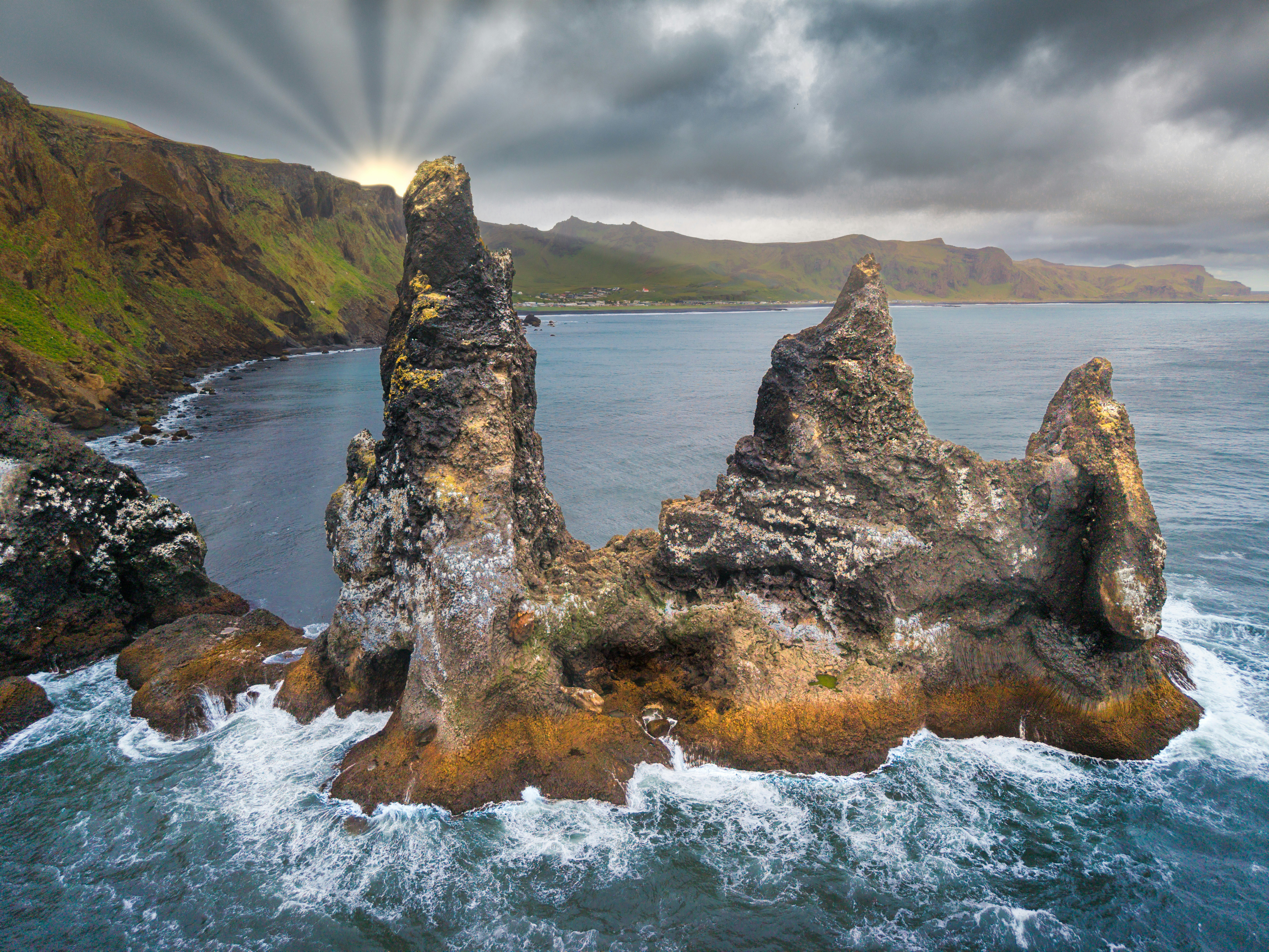 Sunbeams break through storm clouds over the jagged sea stacks of Reynisfjall, with North Atlantic surf crashing against Iceland’s dramatic coastline.