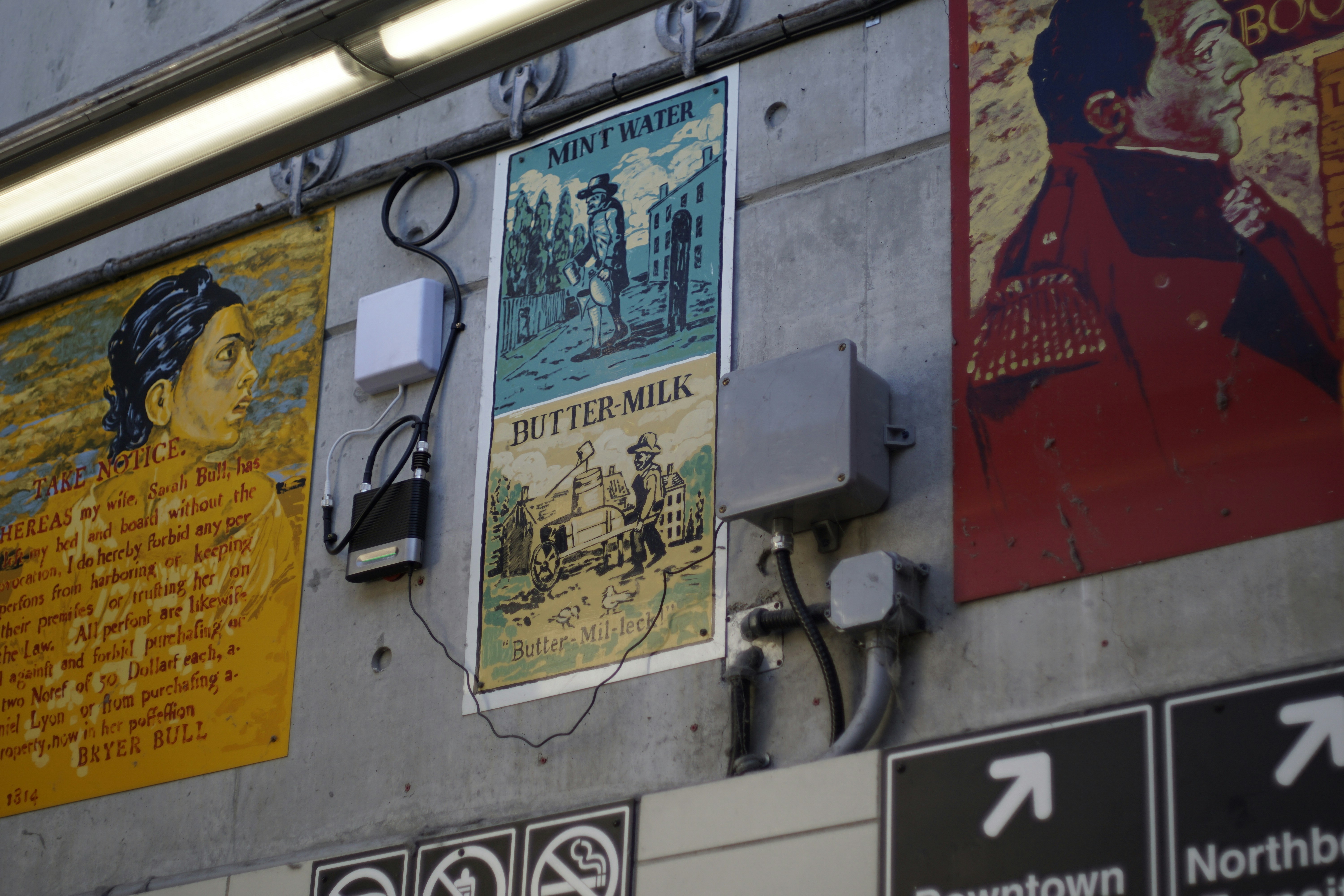Colorful vintage posters mounted on a concrete wall, with industrial conduits and signage beneath, suggesting an urban, retro-factory aesthetic.