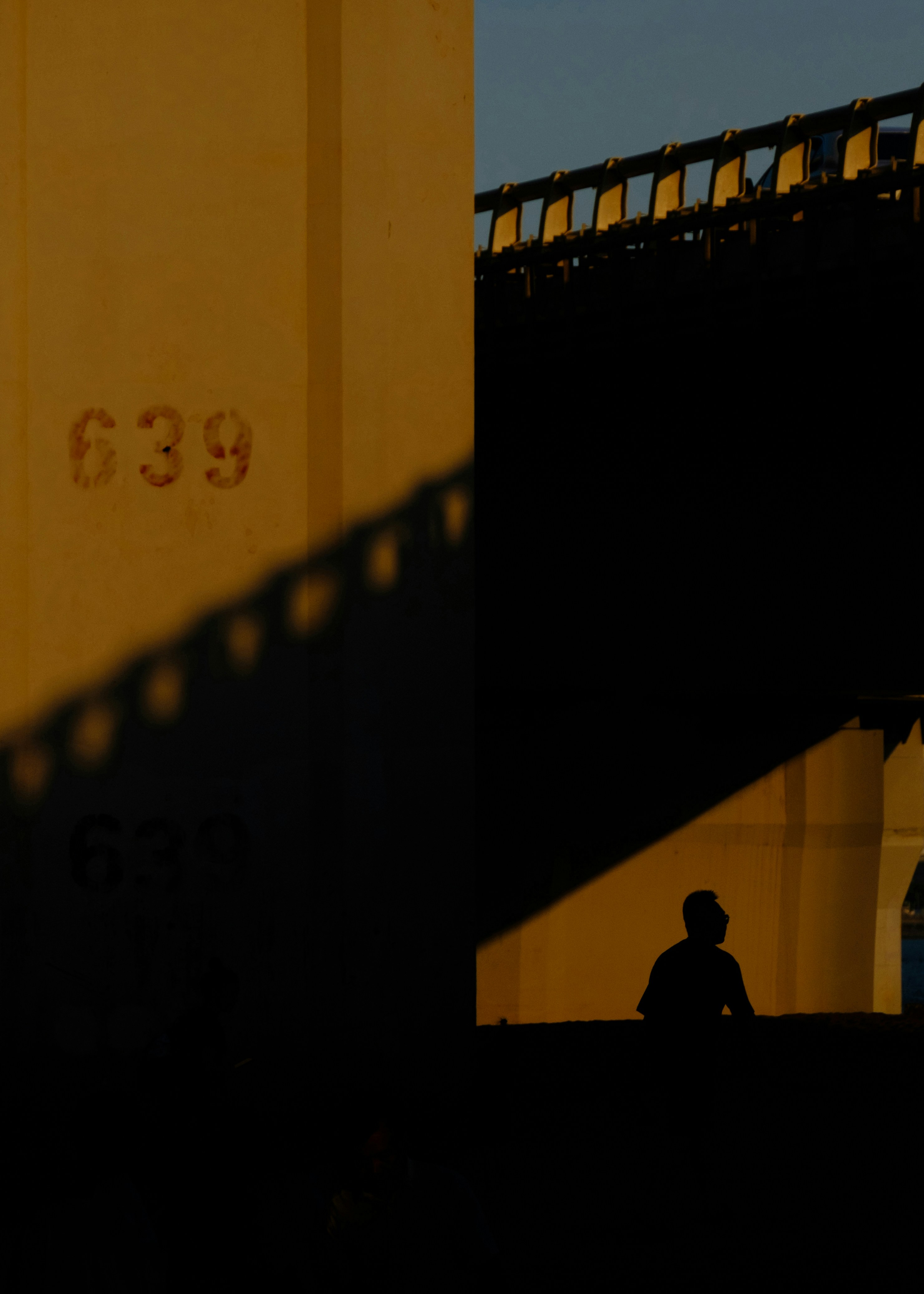  shadowed figure beneath a rusted bridge at golden hour