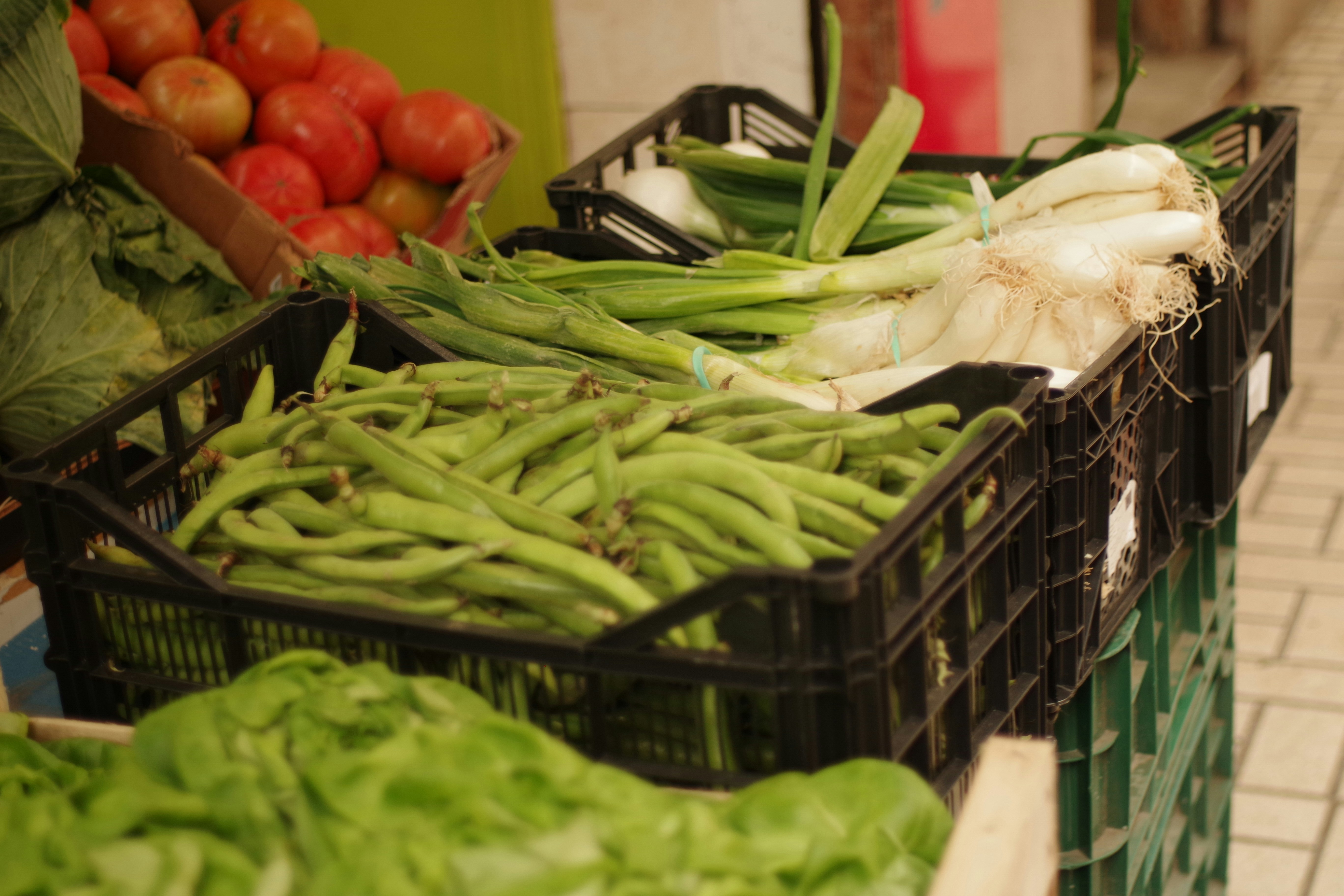 crates of fresh greens line a market stall, onions and spring peas stacked in black crates