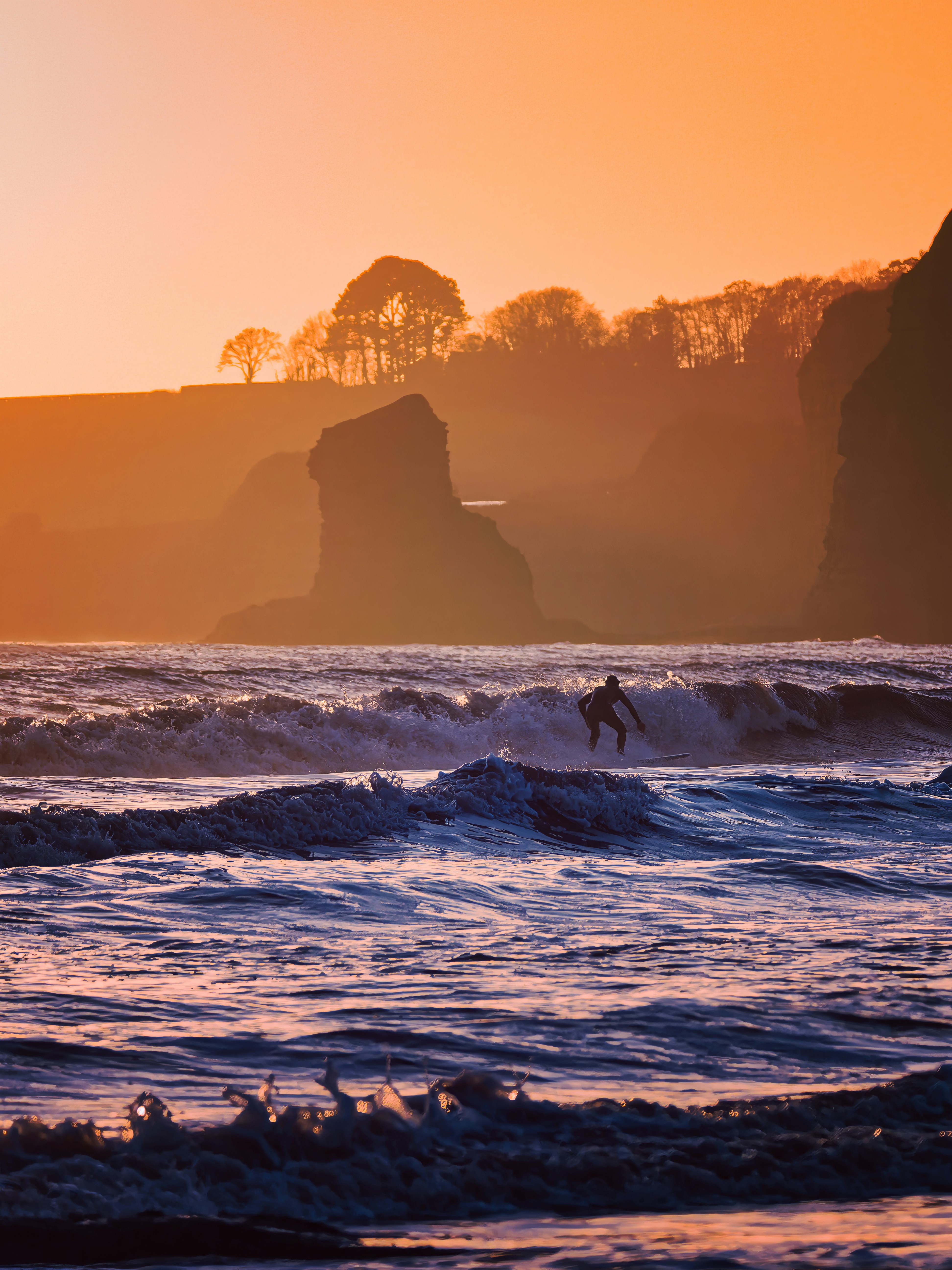 Shadow of the Colossus. Surfer riding the winter high tide with a sunset backdrop of the rock stacks along the Jurassic Coast, Devon, UK