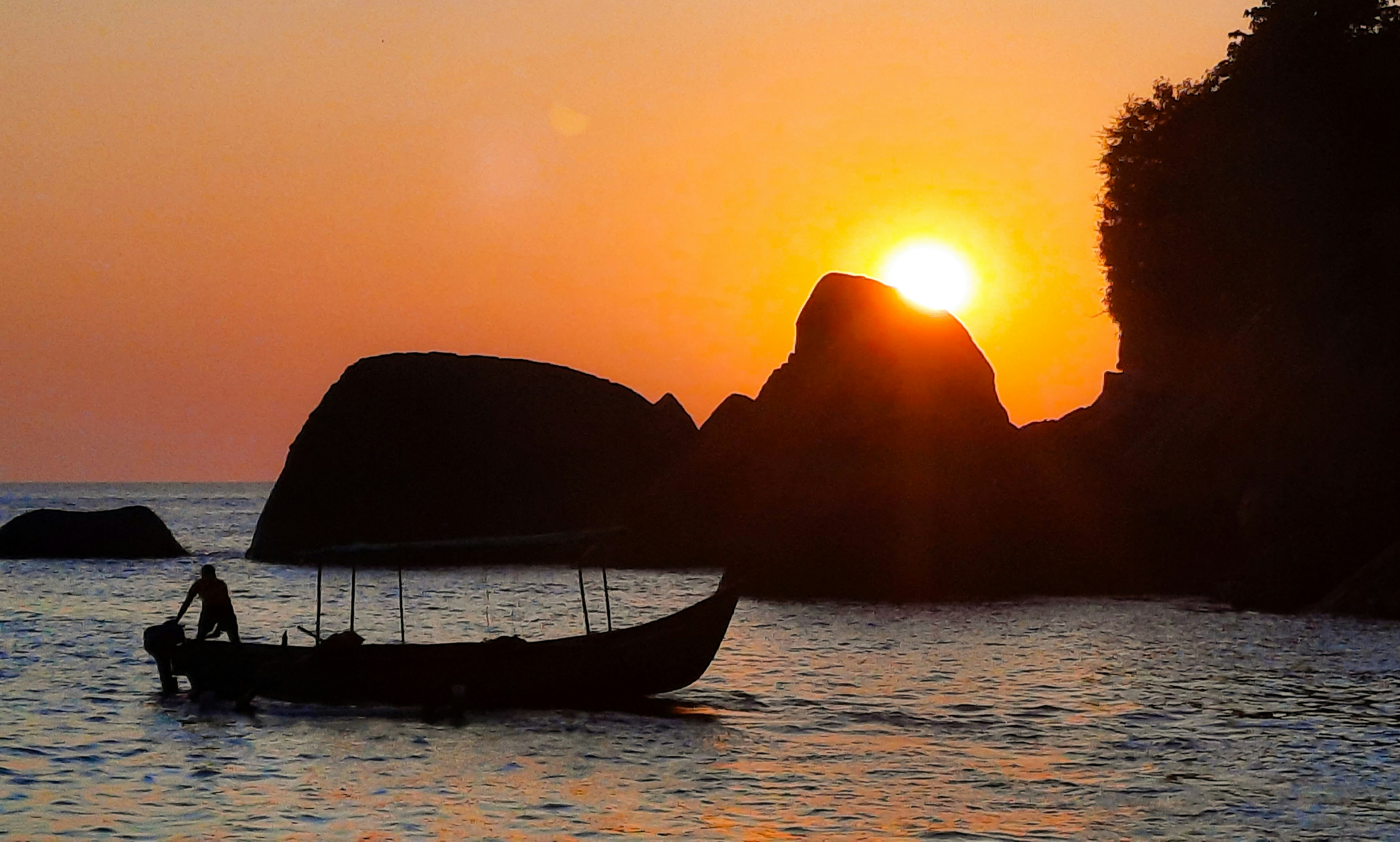 Silhouette of a boat sailing at sunset near rocks.