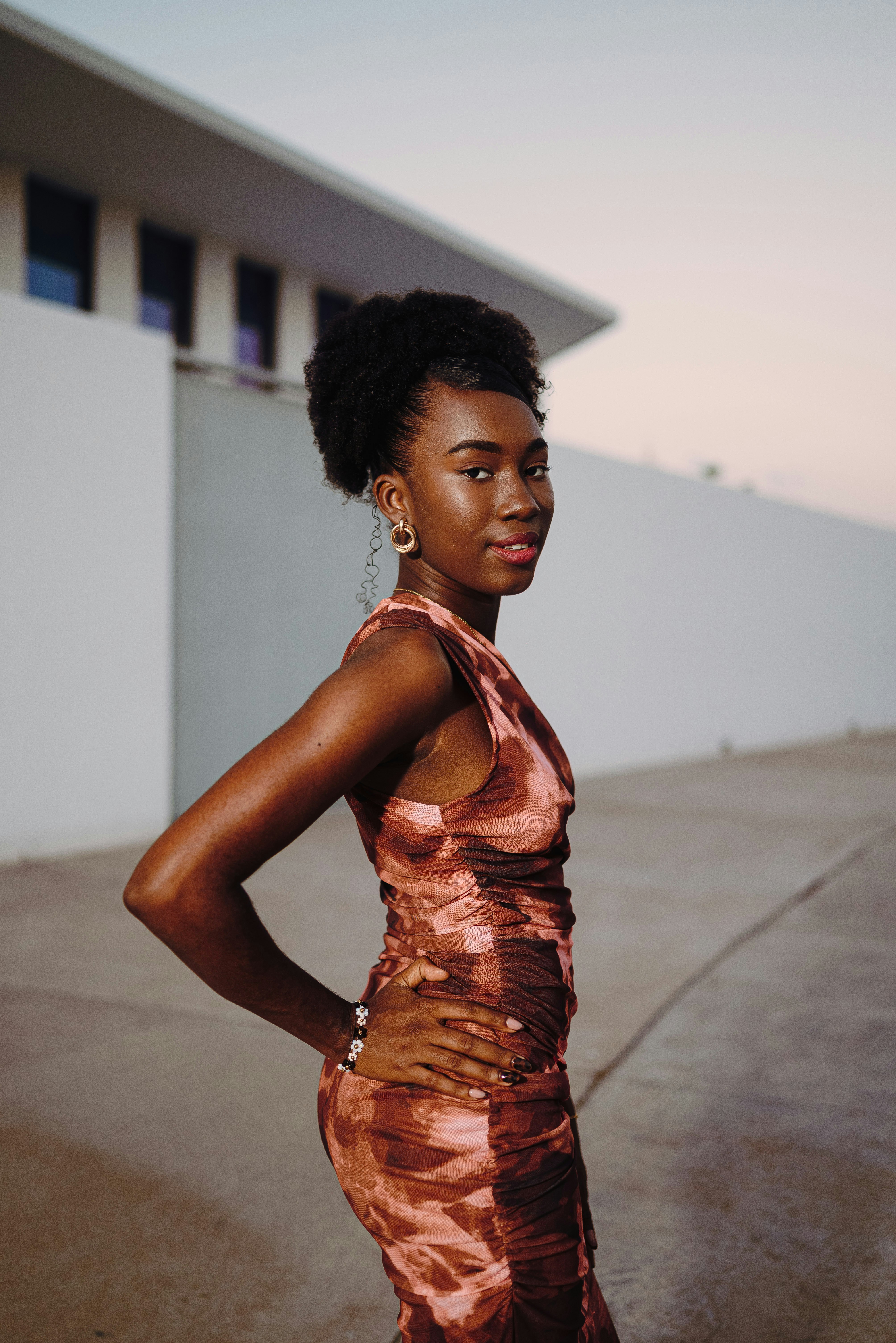 Female model stands in profile against a modern exterior, wearing a copper-toned satin dress; the scene emphasizes silhouette, skin tone, and soft evening light. This is a fashion portrait photo.