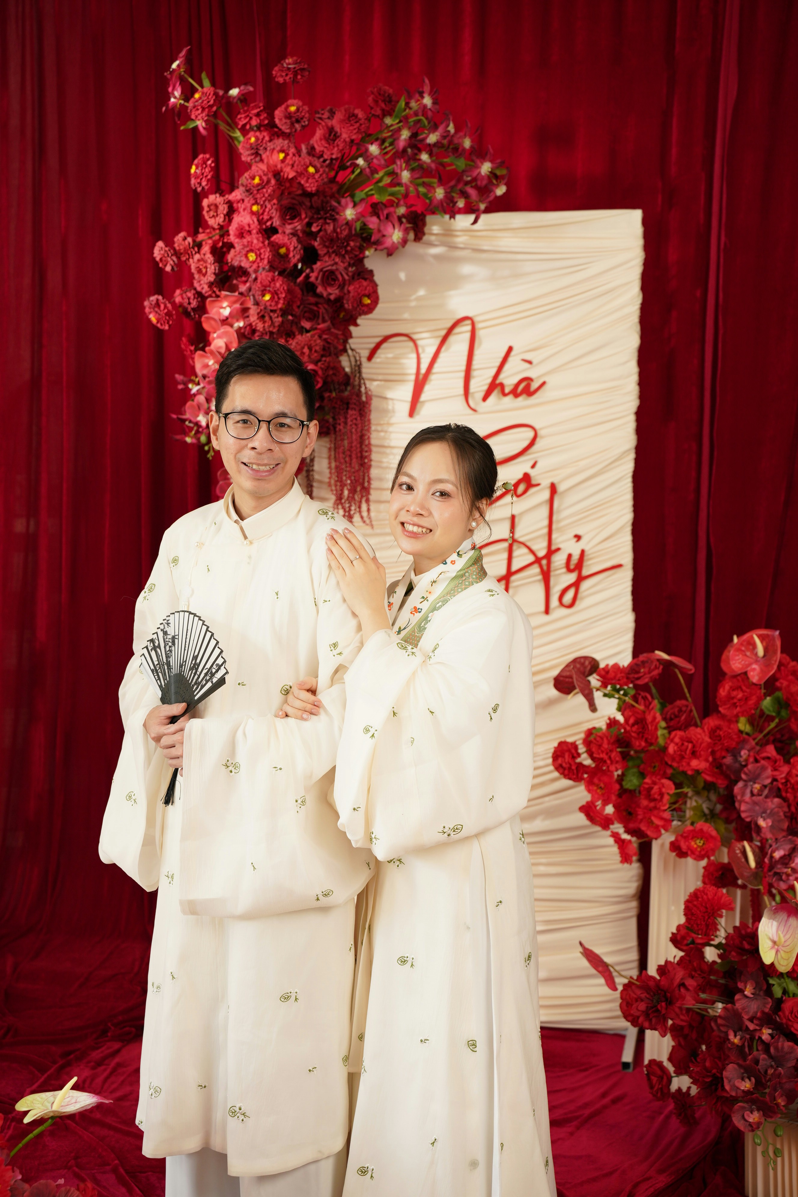 Elegance in White: A Couple in Traditional Attire Amid Red Floral Backdrop