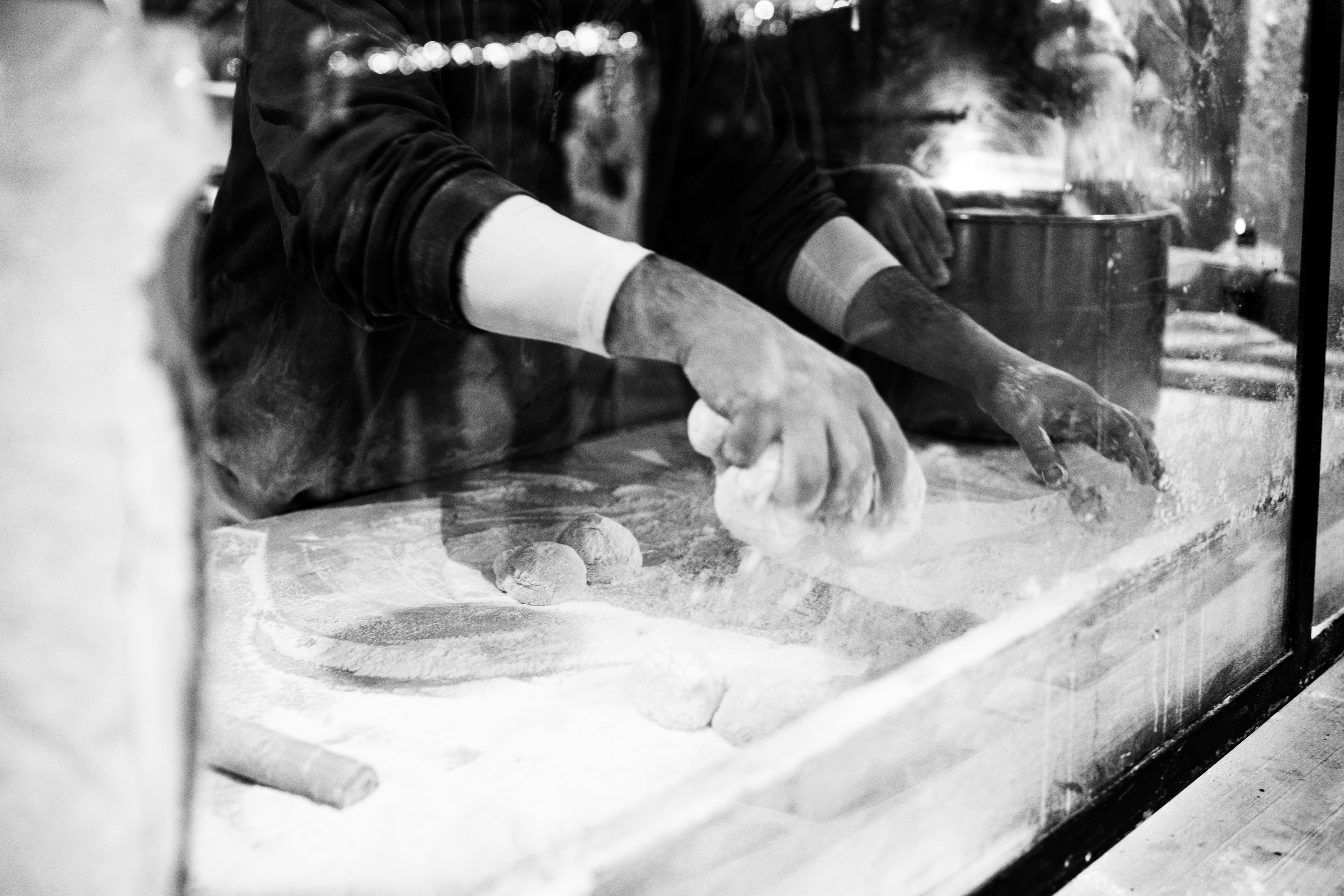 A monochrome bakery scene shows hands shaping dough inside a glass display, with reflections and flour dust creating soft textures. The focus is on the artisan activity and the tactile process of bread making.