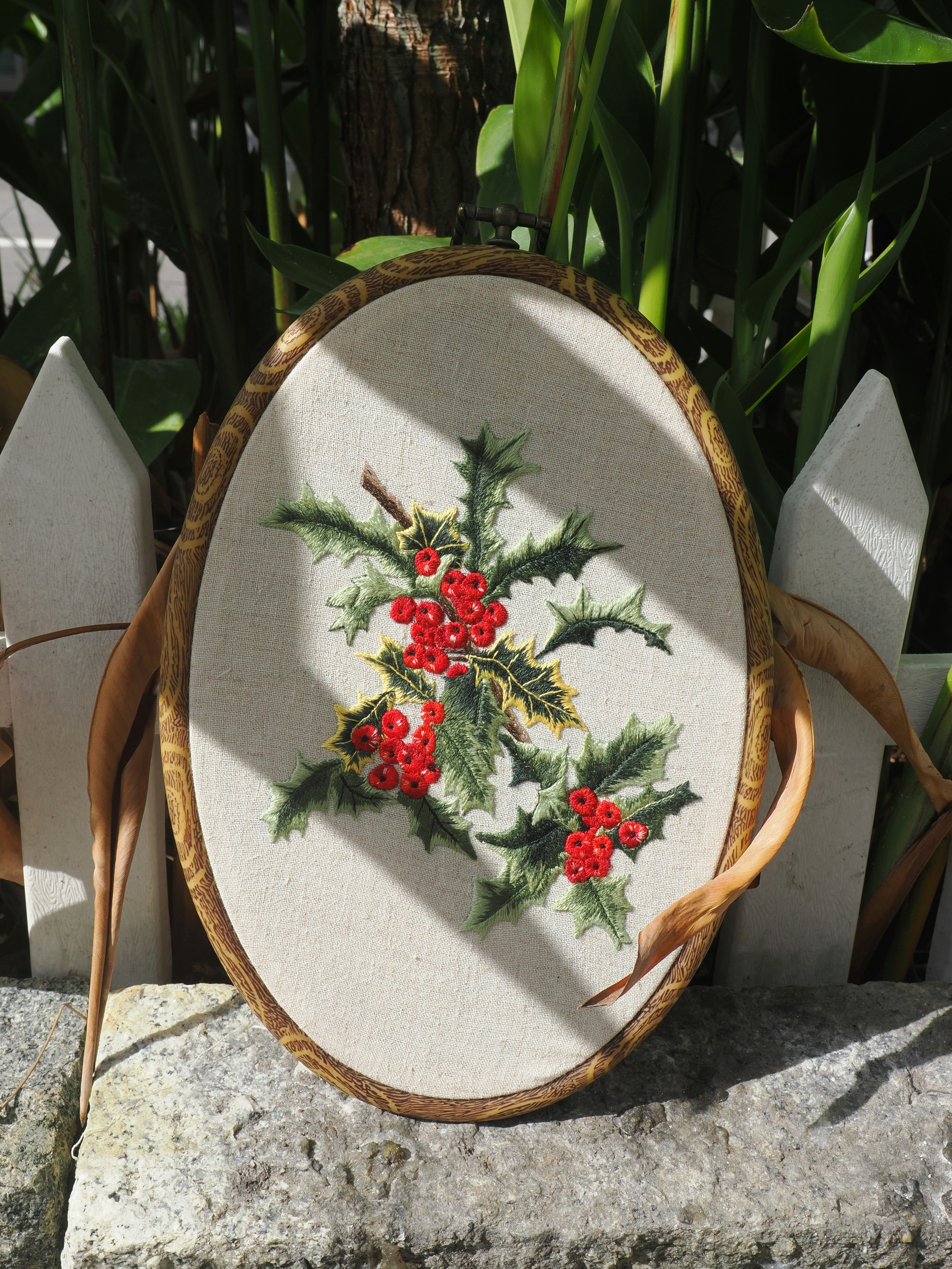 Embroidery hoop with holly and red berries set against a garden backdrop, framed by a wooden hoop and leaning on a rustic stone surface.