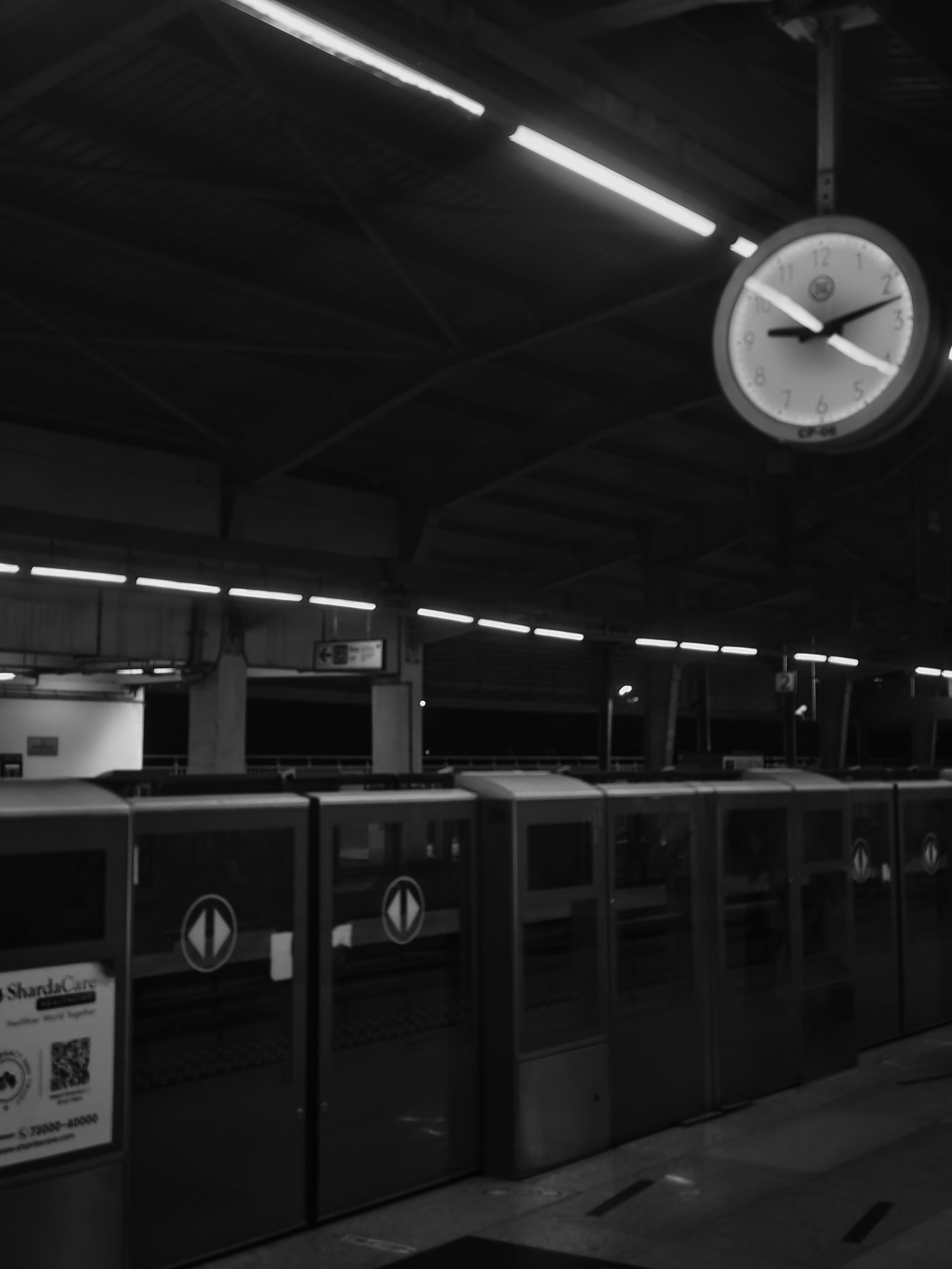 A moody, high-contrast black and white shot of a station clock at night. The clock is framed by sharp, linear light fixtures, emphasizing the urban atmosphere and the quiet stillness of a transit platform after hours
