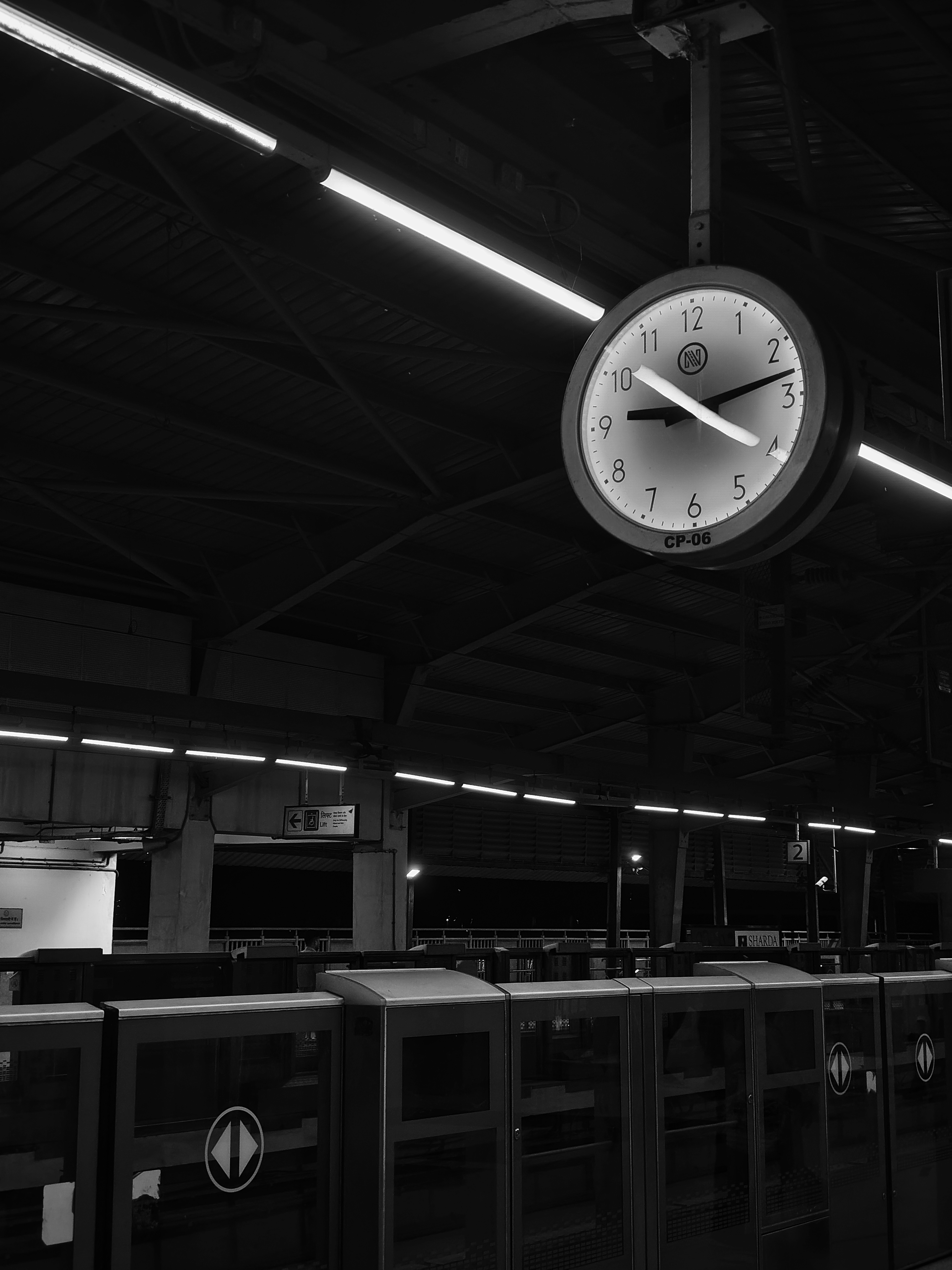 A moody, high-contrast black and white shot of a station clock at night. The clock is framed by sharp, linear light fixtures, emphasizing the urban atmosphere and the quiet stillness of a transit platform after hours