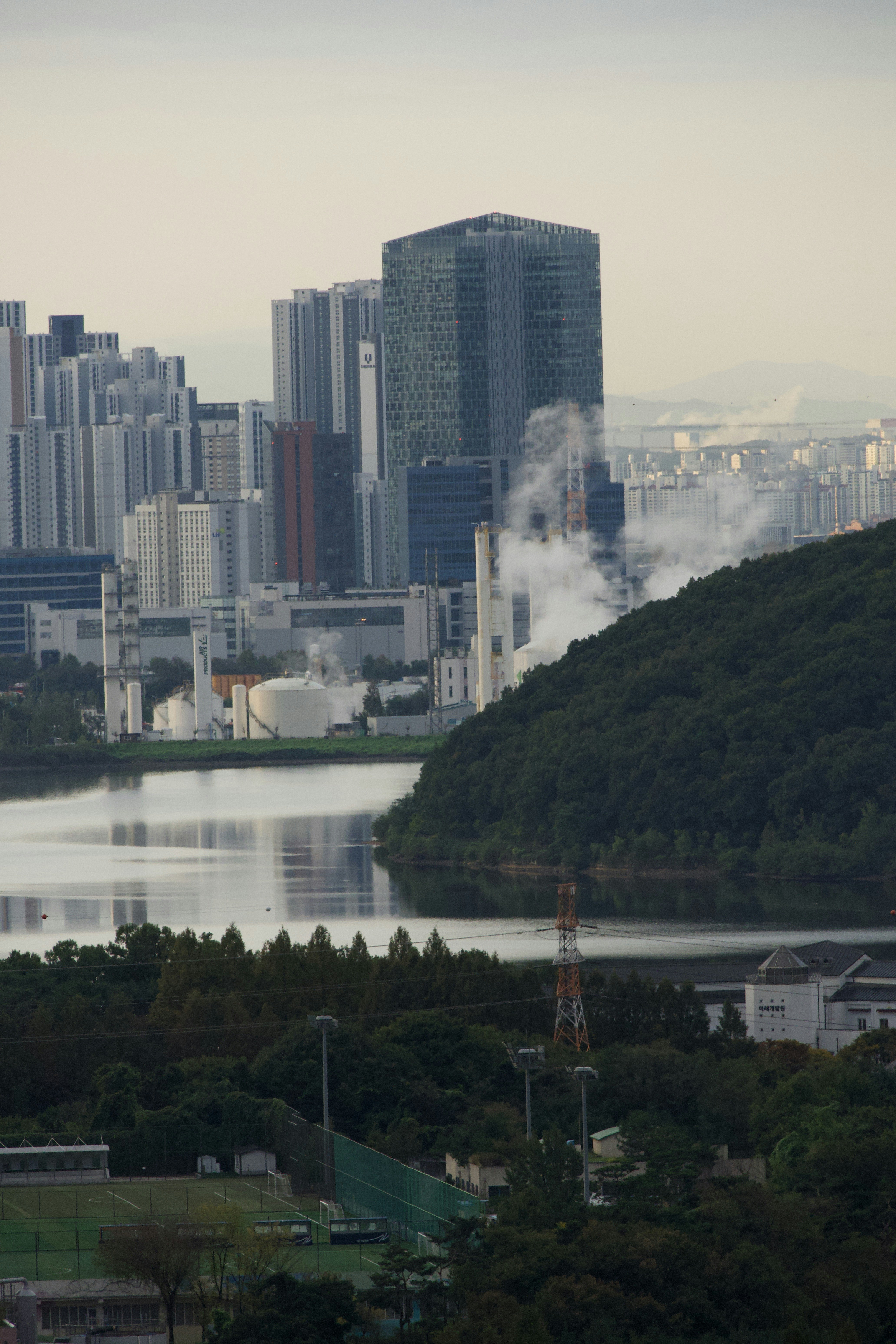 Vertical Cityscape: Towers, Smoke, and a Green Ridge by the Water