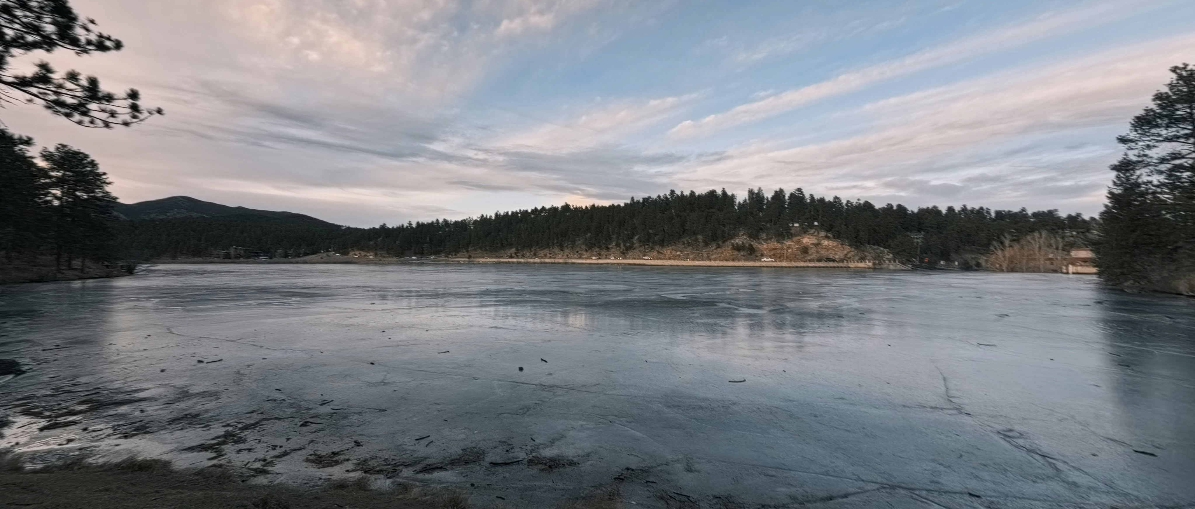 Frozen lake surrounded by pine trees under cloudy sky.
