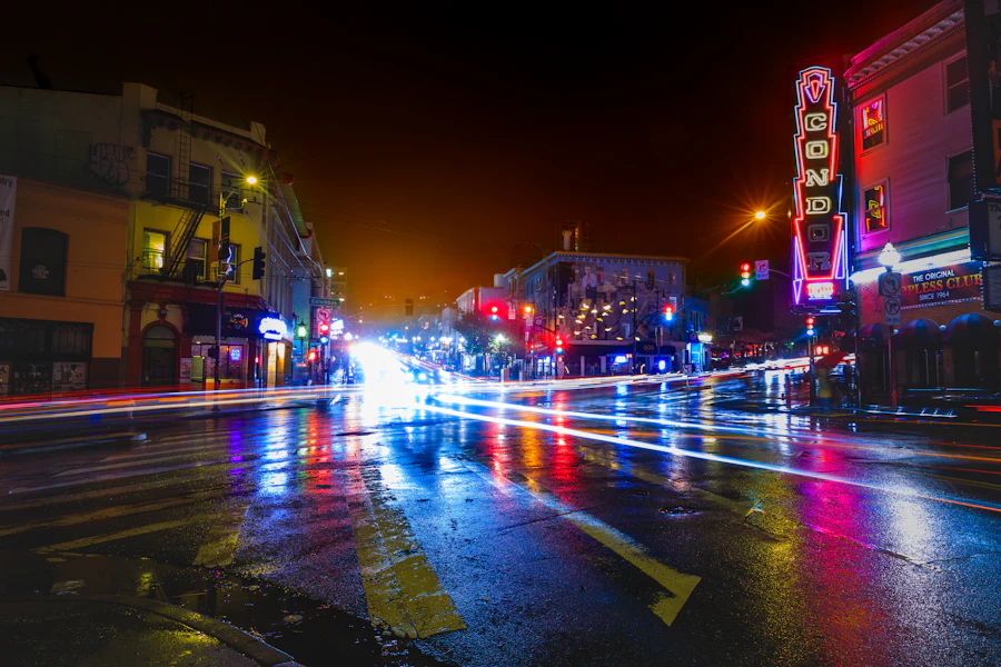 Nighttime city street with neon signs and light trails across a wet intersection, capturing motion and reflections in the scene.
