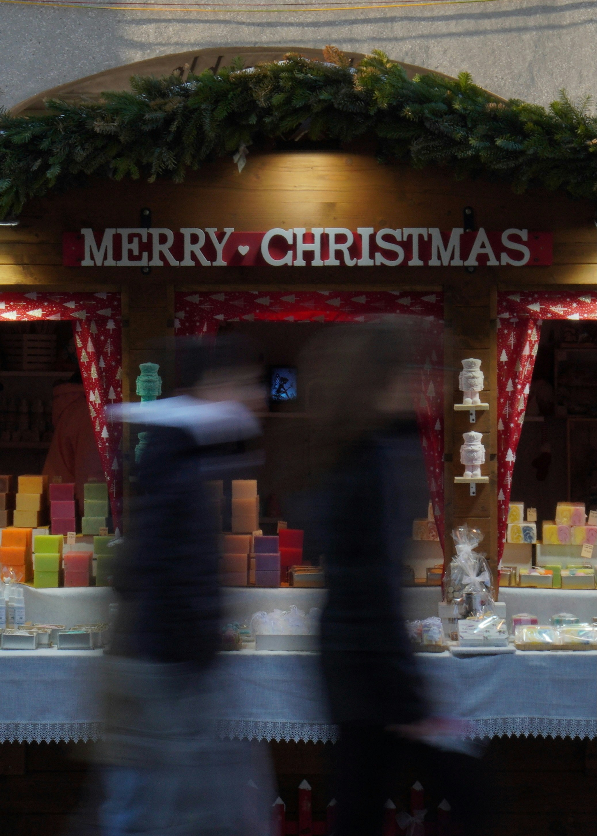 Blurred figures pass in front of a festive Christmas market stall beneath a wooden storefront and evergreen garland with a bright 'MERRY CHRISTMAS' sign.