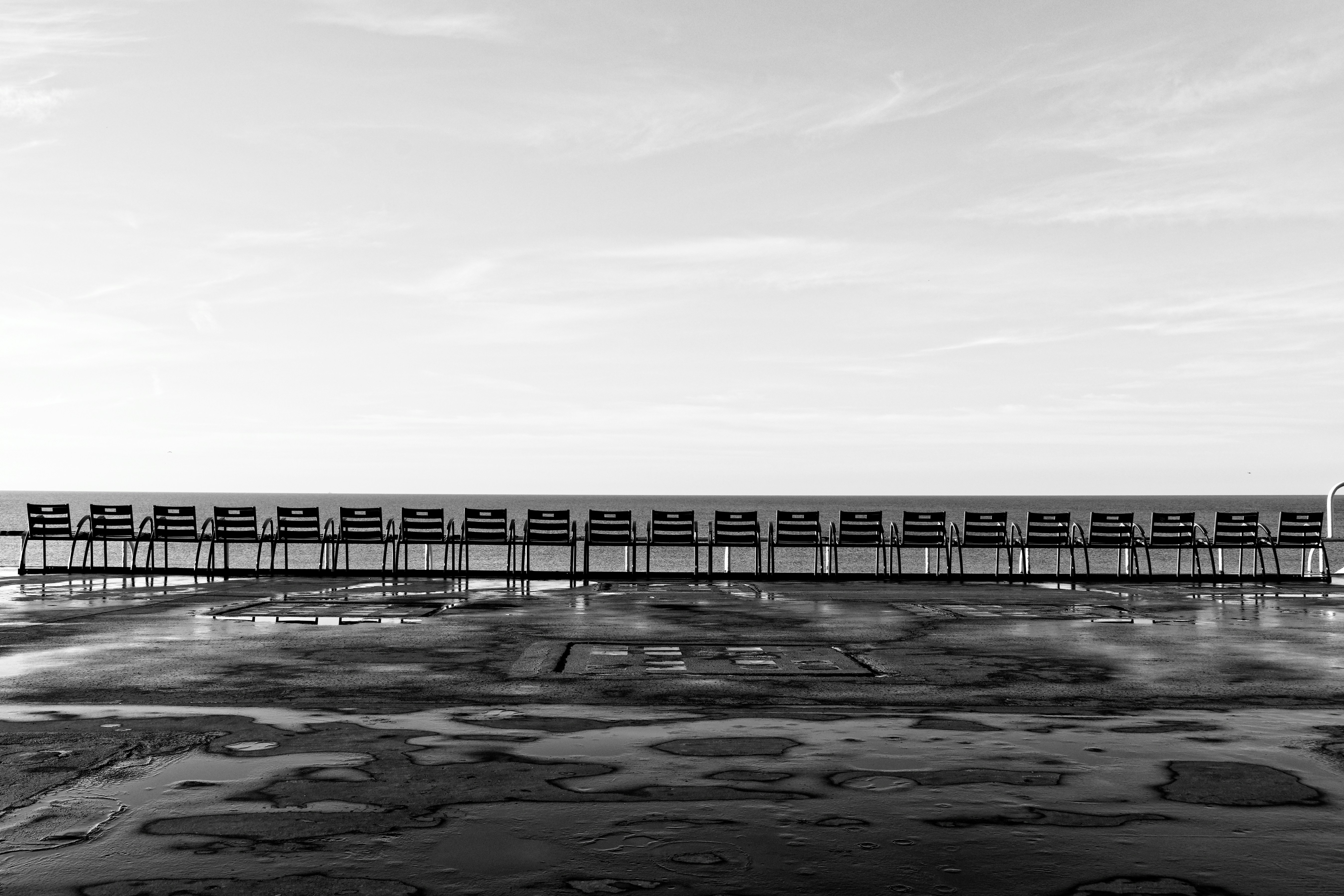 Des chaises vides alignées sur la Promenade des Anglais à Nice créent une composition minimaliste et contemplative. Cette perspective en noir et blanc souligne la géométrie et la sérénité matinale sur la Prom. Offrant une vision poétique de la vie côtière méditerranéenne au repos.
