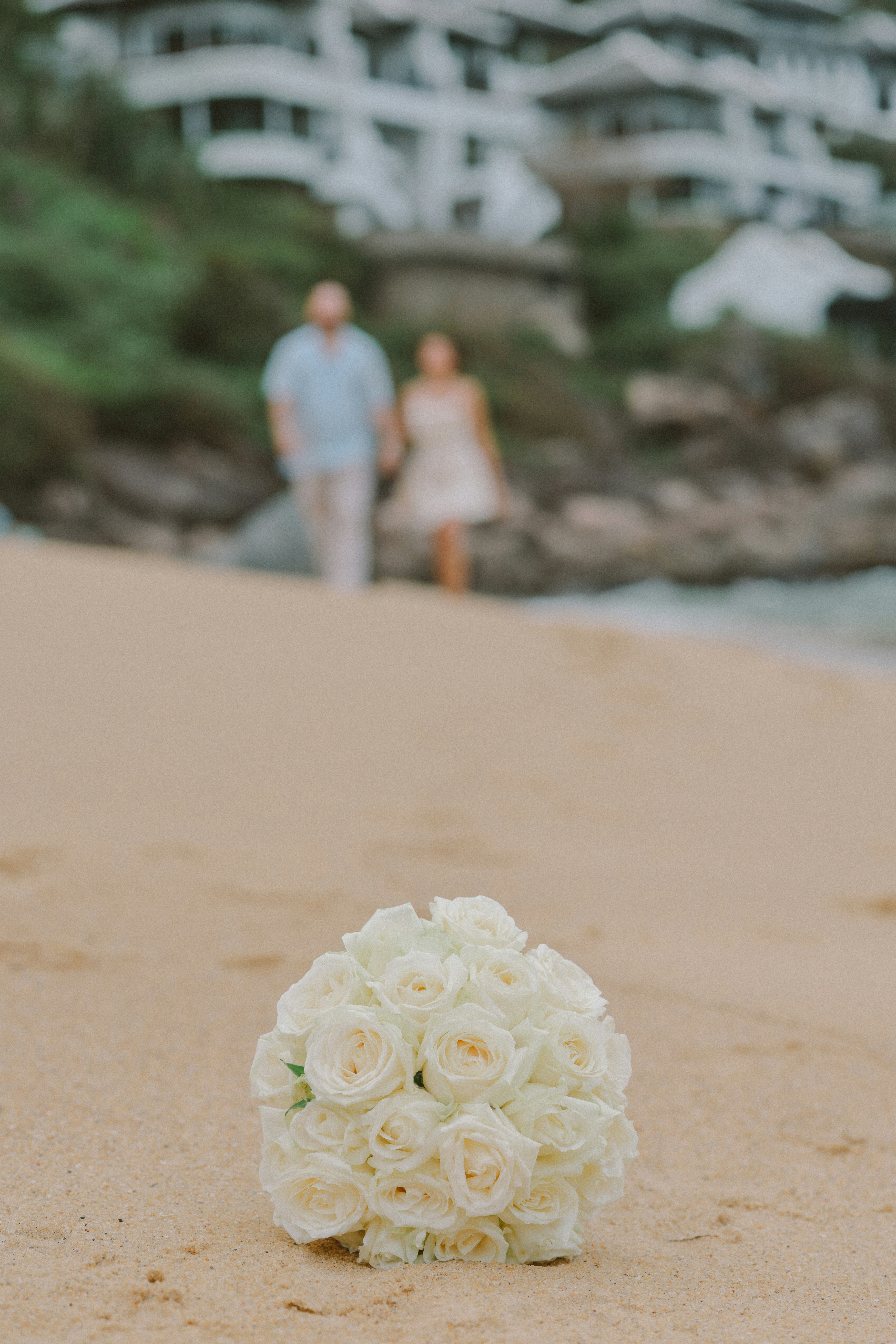White bouquet on sunlit sand with blurred couple and resort backdrop