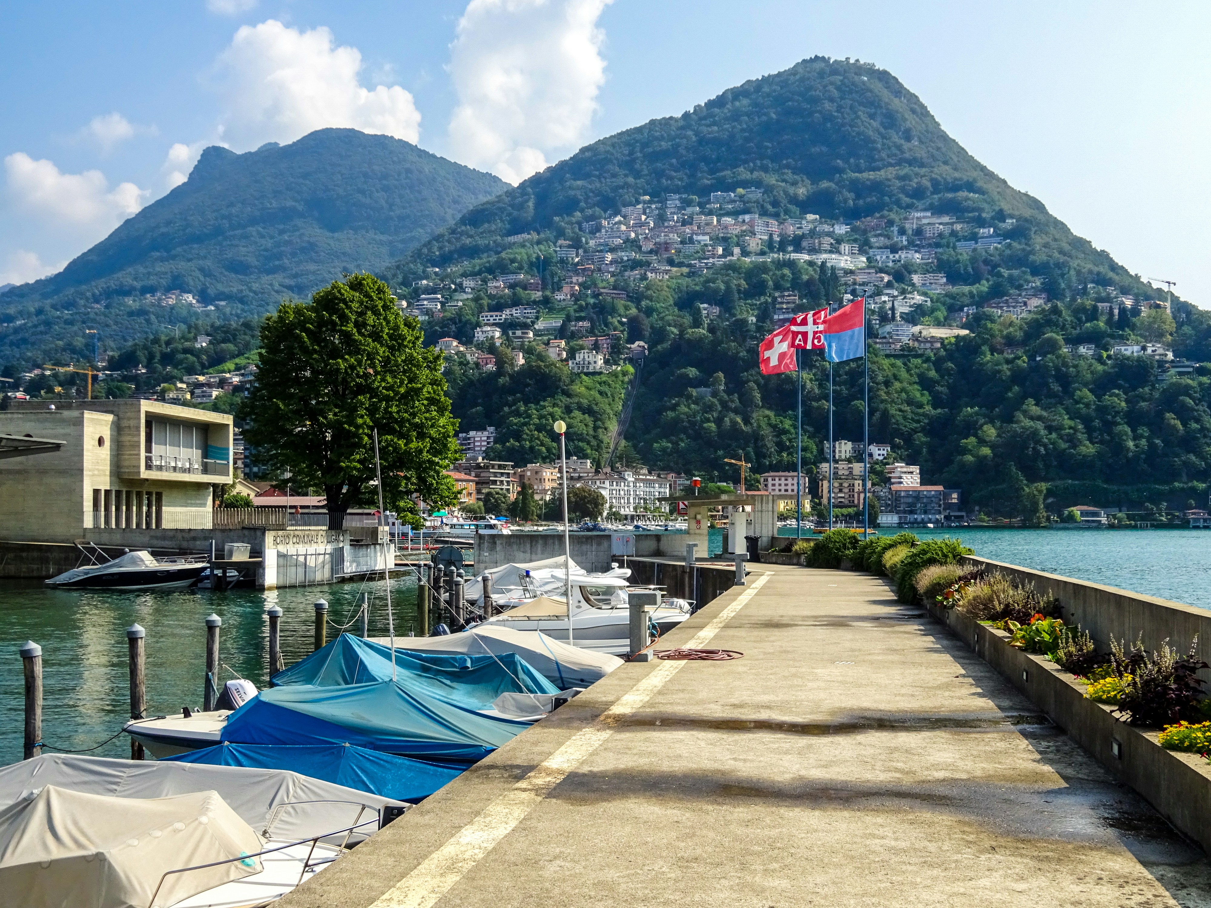 Boats docked by a walkway with mountains and buildings.