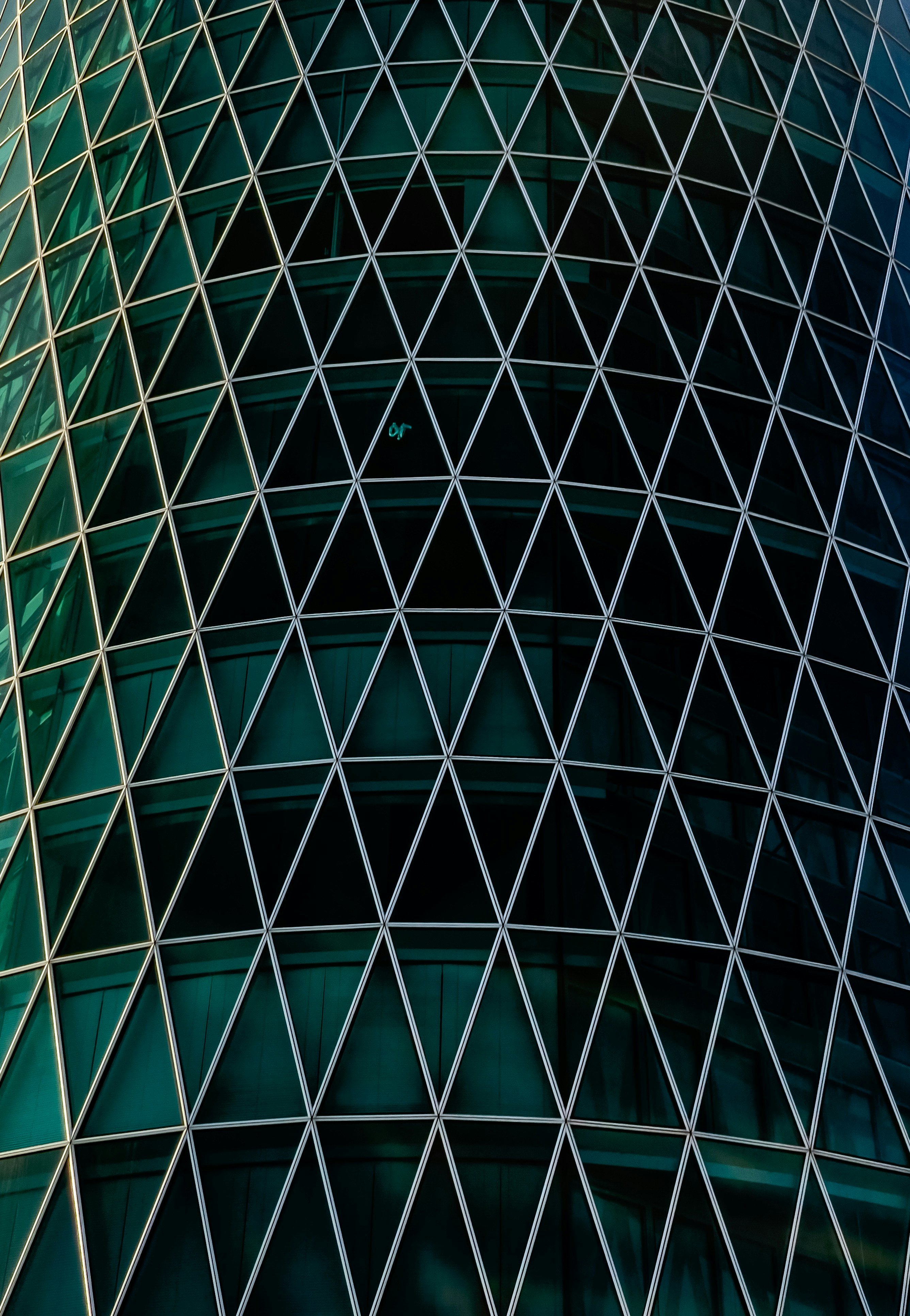 Close-up of a diamond-pattern glass facade with a dark teal and green gradient, forming a repetitive lattice across the curved surface.