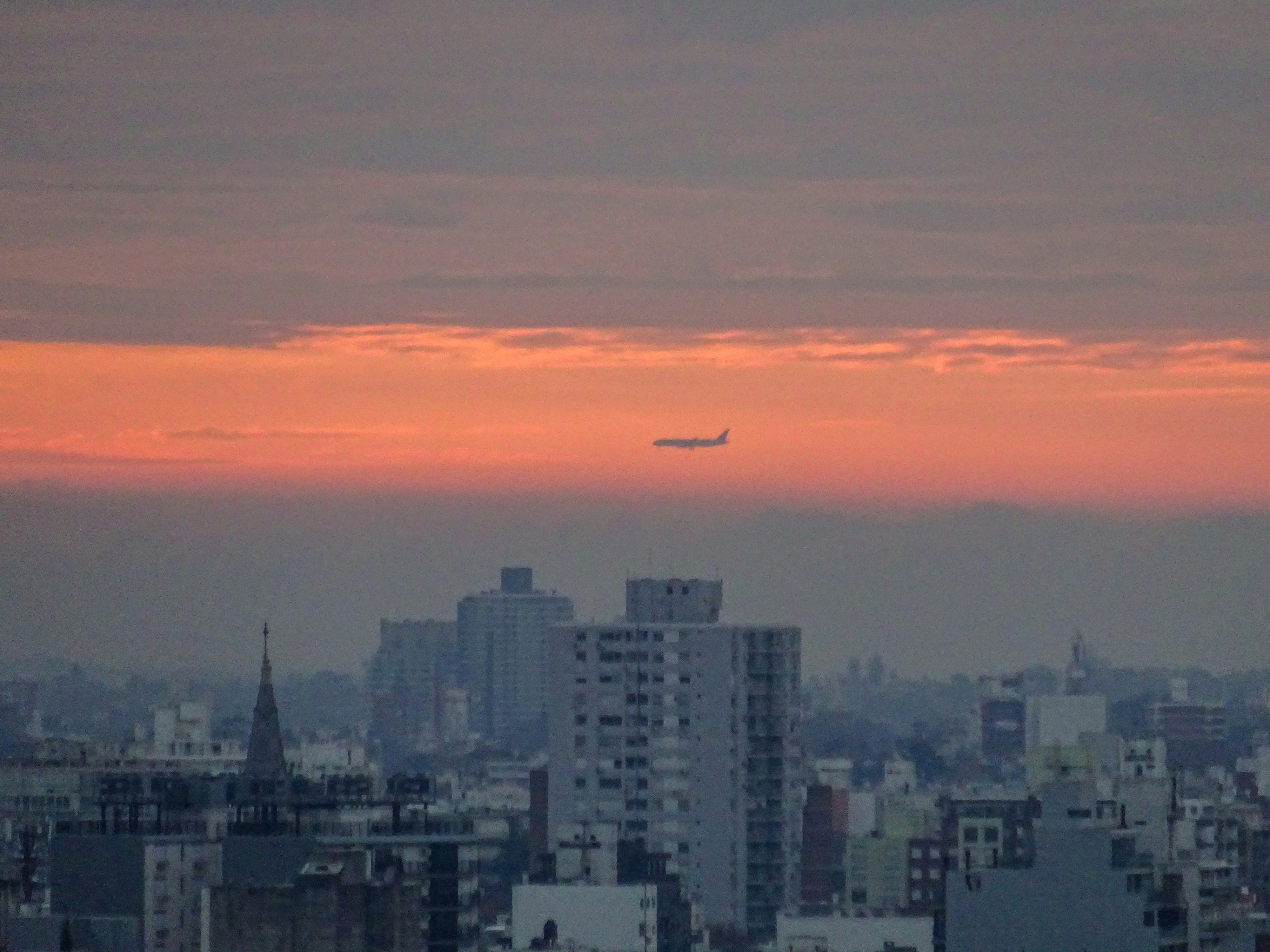 Airplane flies over city buildings at sunset.