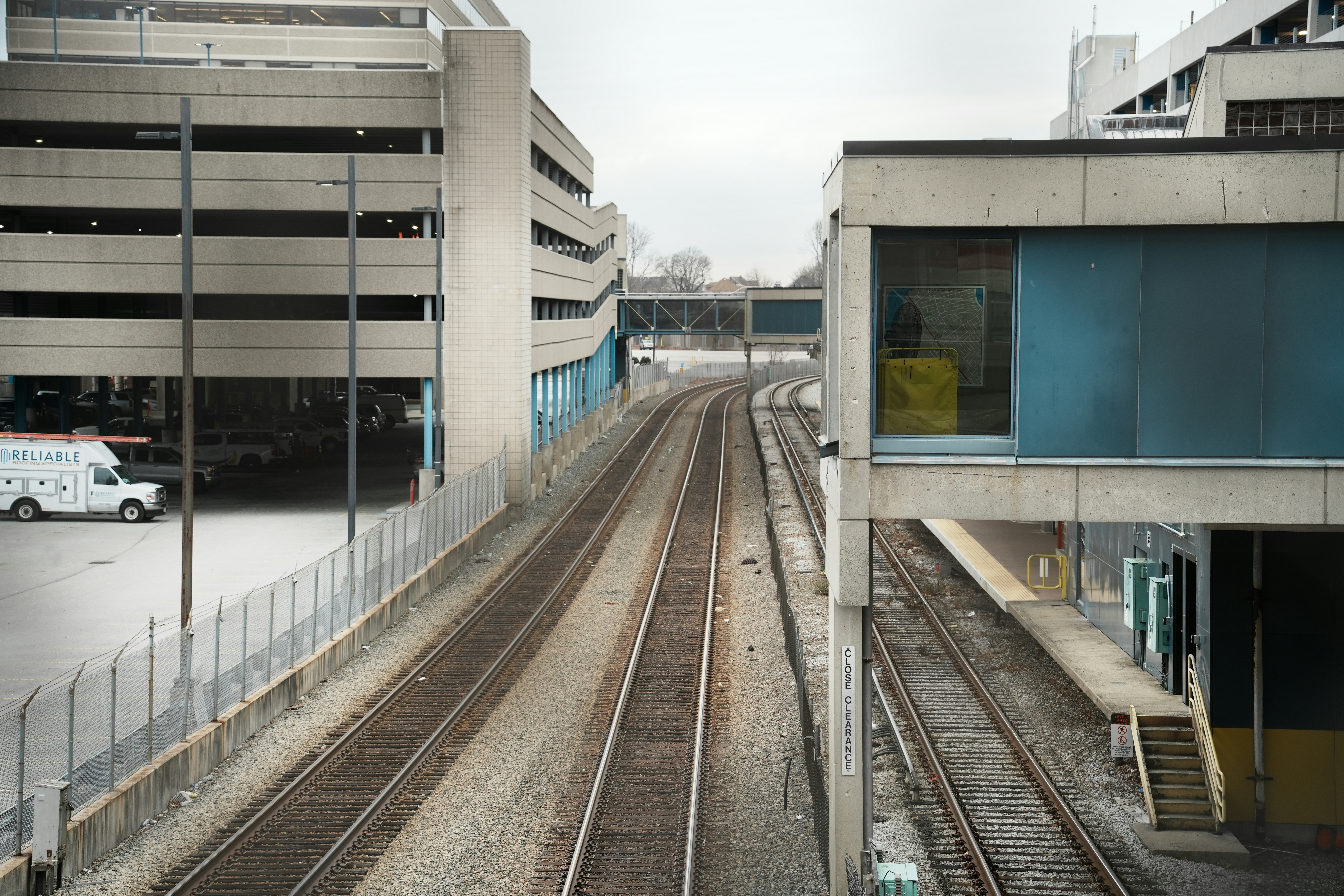 Railway tracks converge beneath a concrete overpass, with a distant vanishing point between two station structures. The scene emphasizes industrial urban geometry and sparse color.