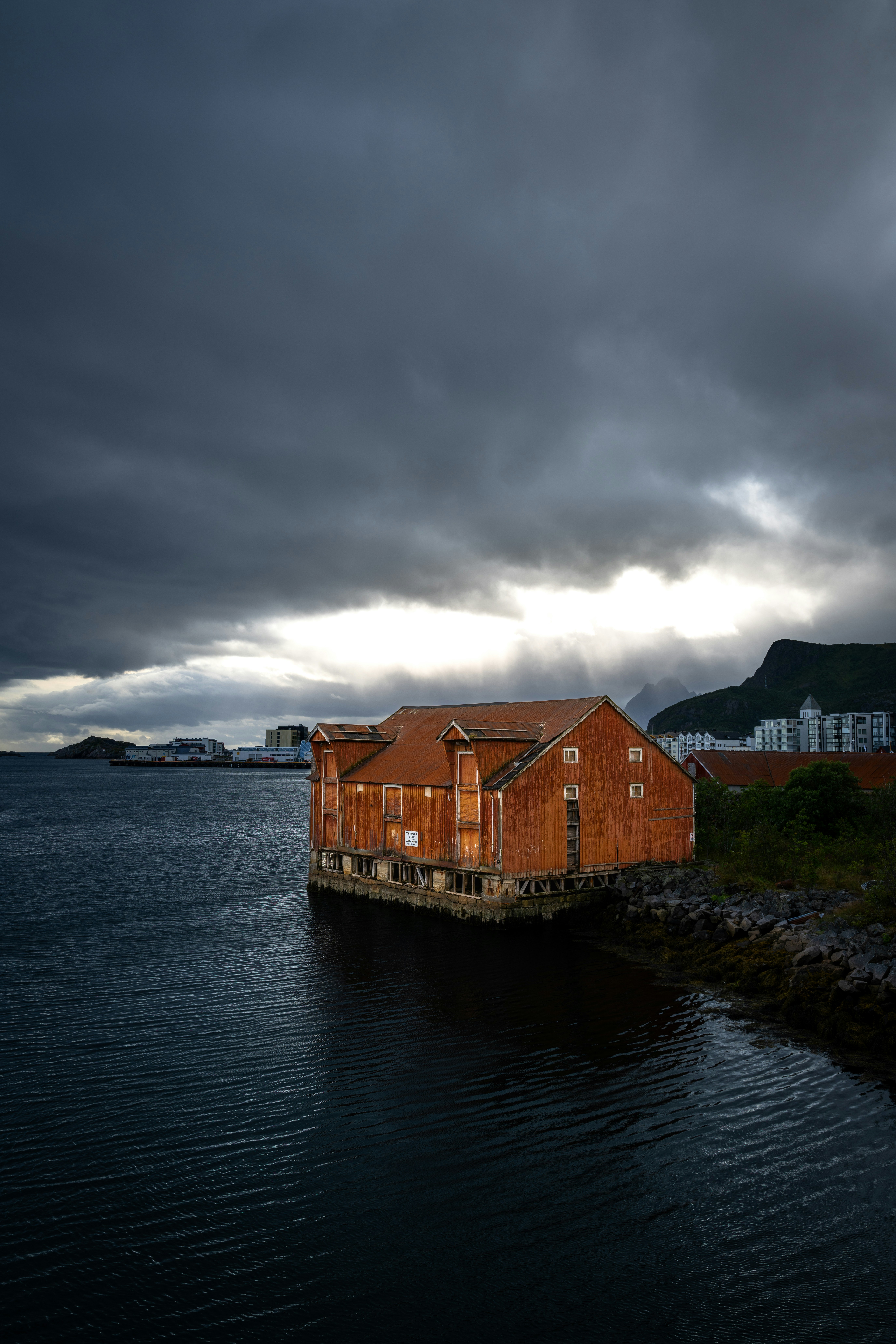 Rust-red boathouse anchored to rocky shore under dramatic skies
