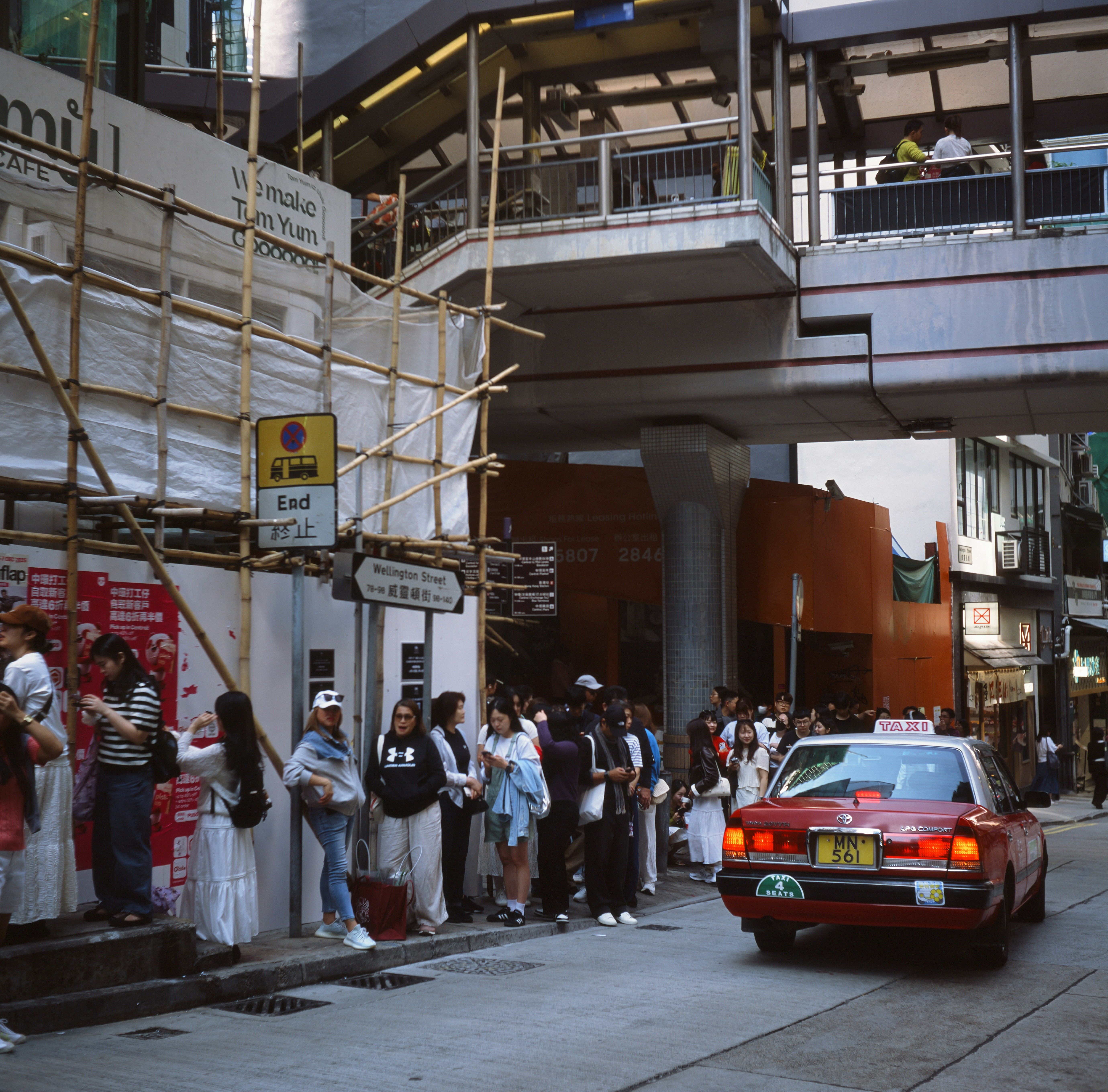 People queueing for transport near a red taxi.