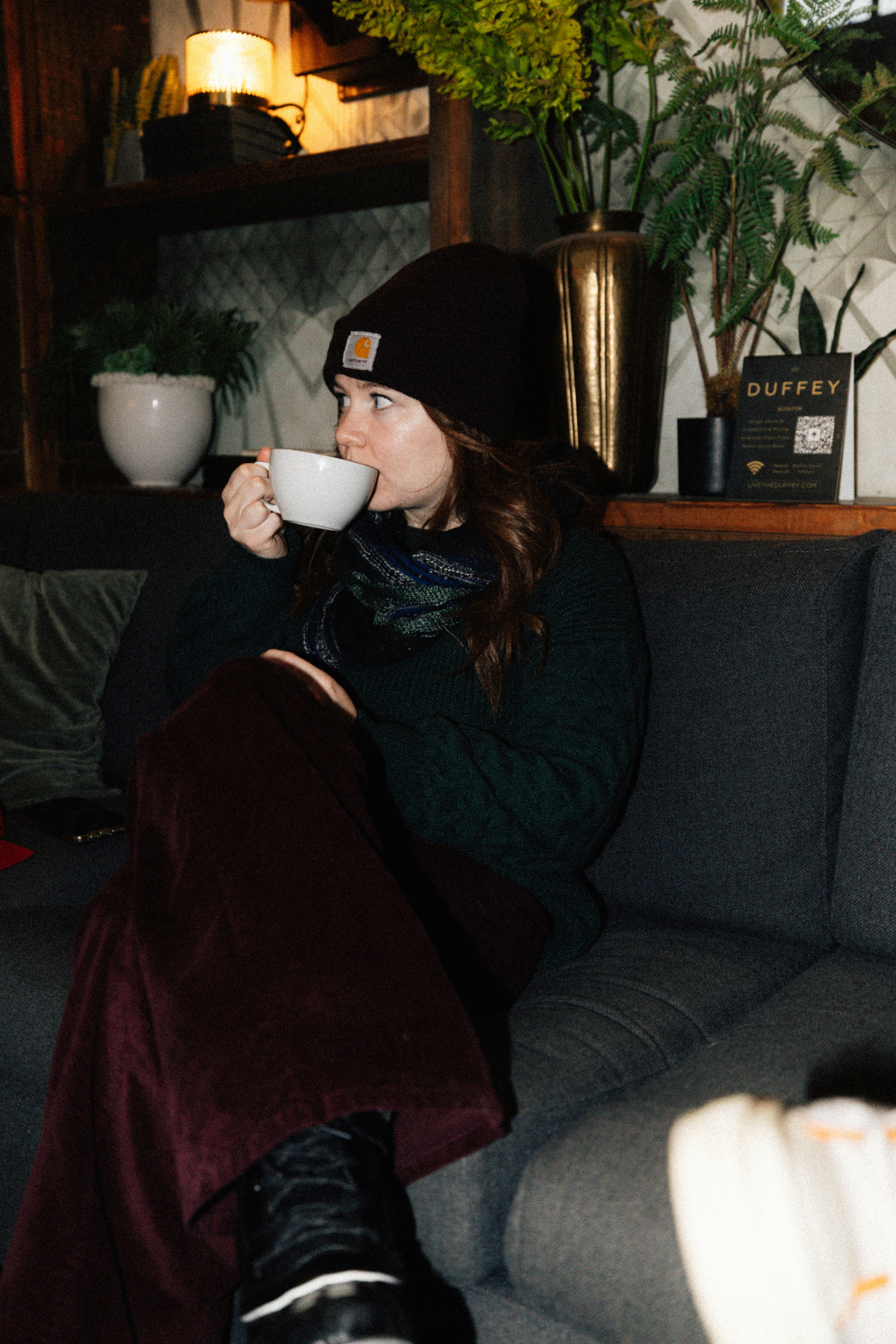 Cozy Quiet Moment: Woman in Knit Hat Sips from a White Cup on a Dark Sofa