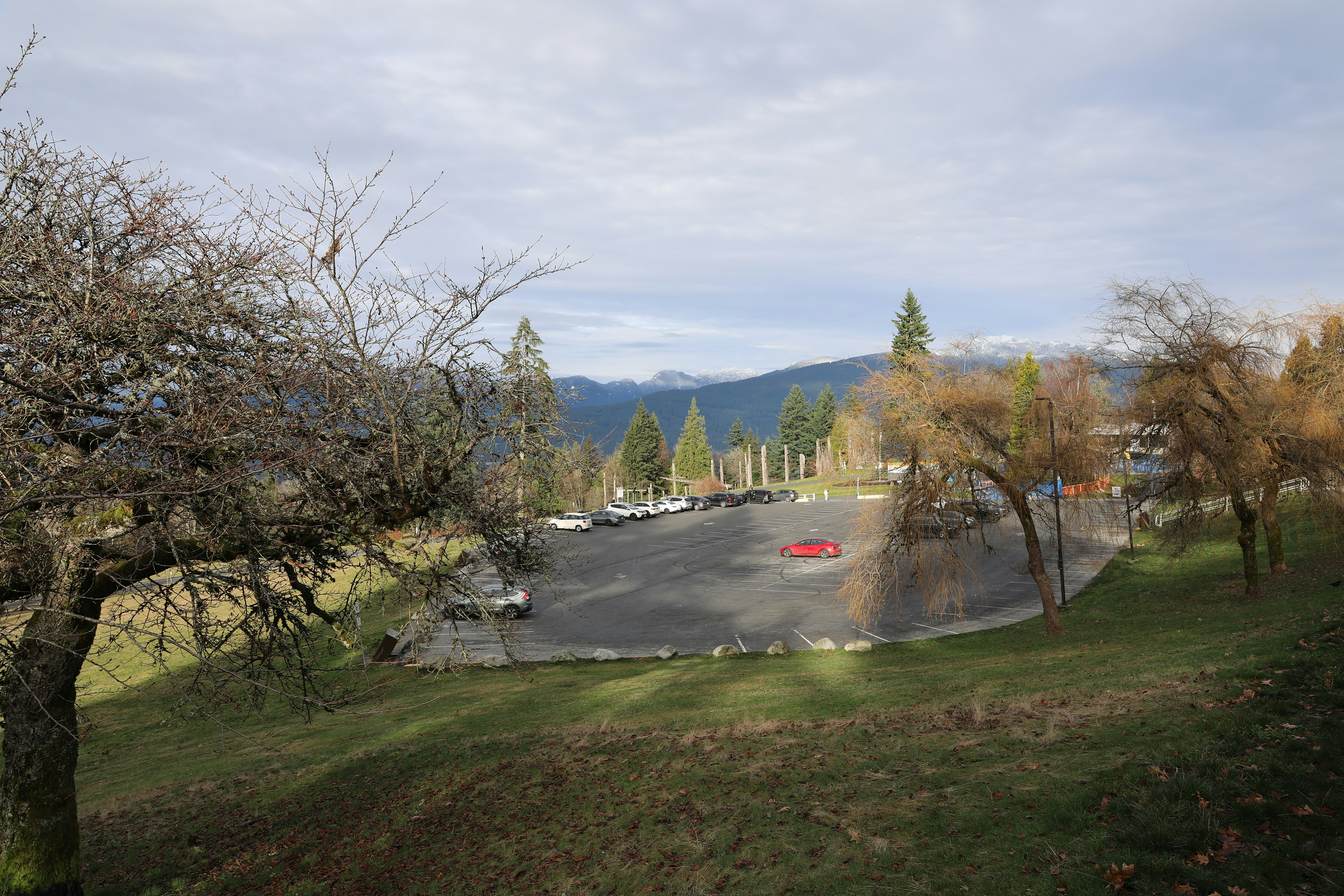 Parking lot with several cars framed by bare trees and a distant mountain range under a cloudy sky. The scene suggests a calm park-like area adjacent to the lot.