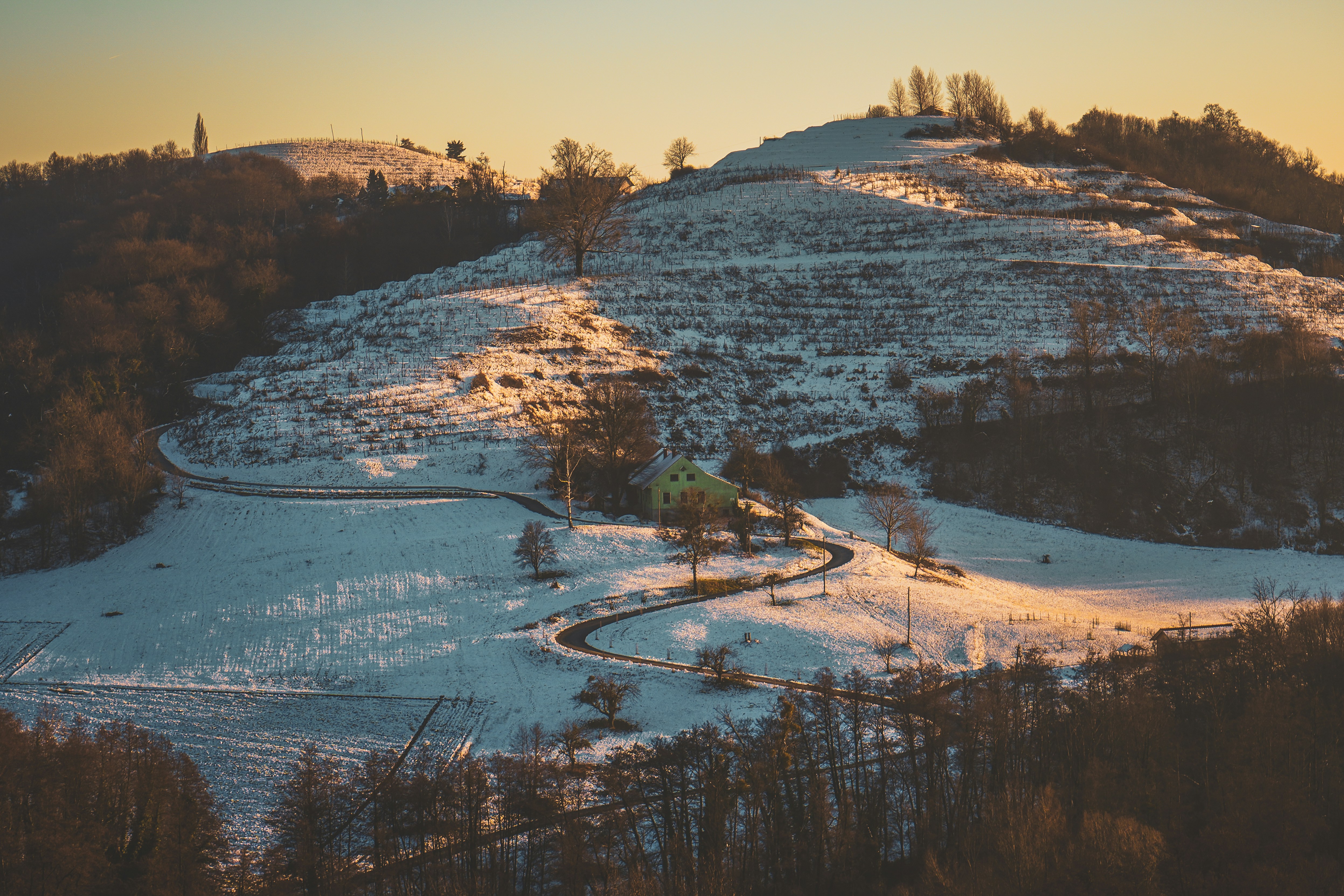 Snow-tinged hillside road meanders toward a quiet green cabin at sunset