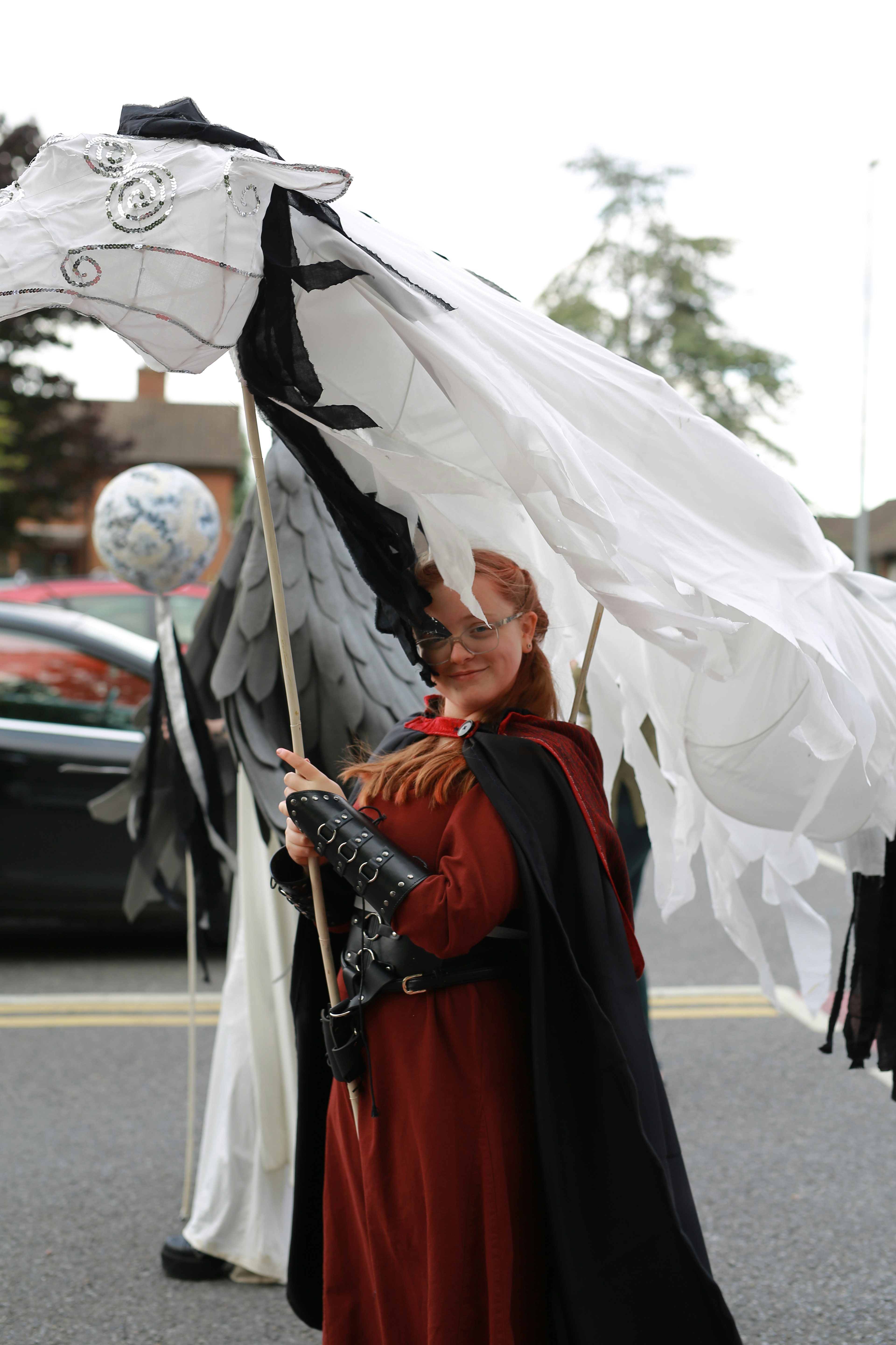 A young girl in a red cloak and black corset stands in a parade, holding a dragon wing prop with white fabric. The scene includes balloons and parked cars in the background, conveying a festive street event.