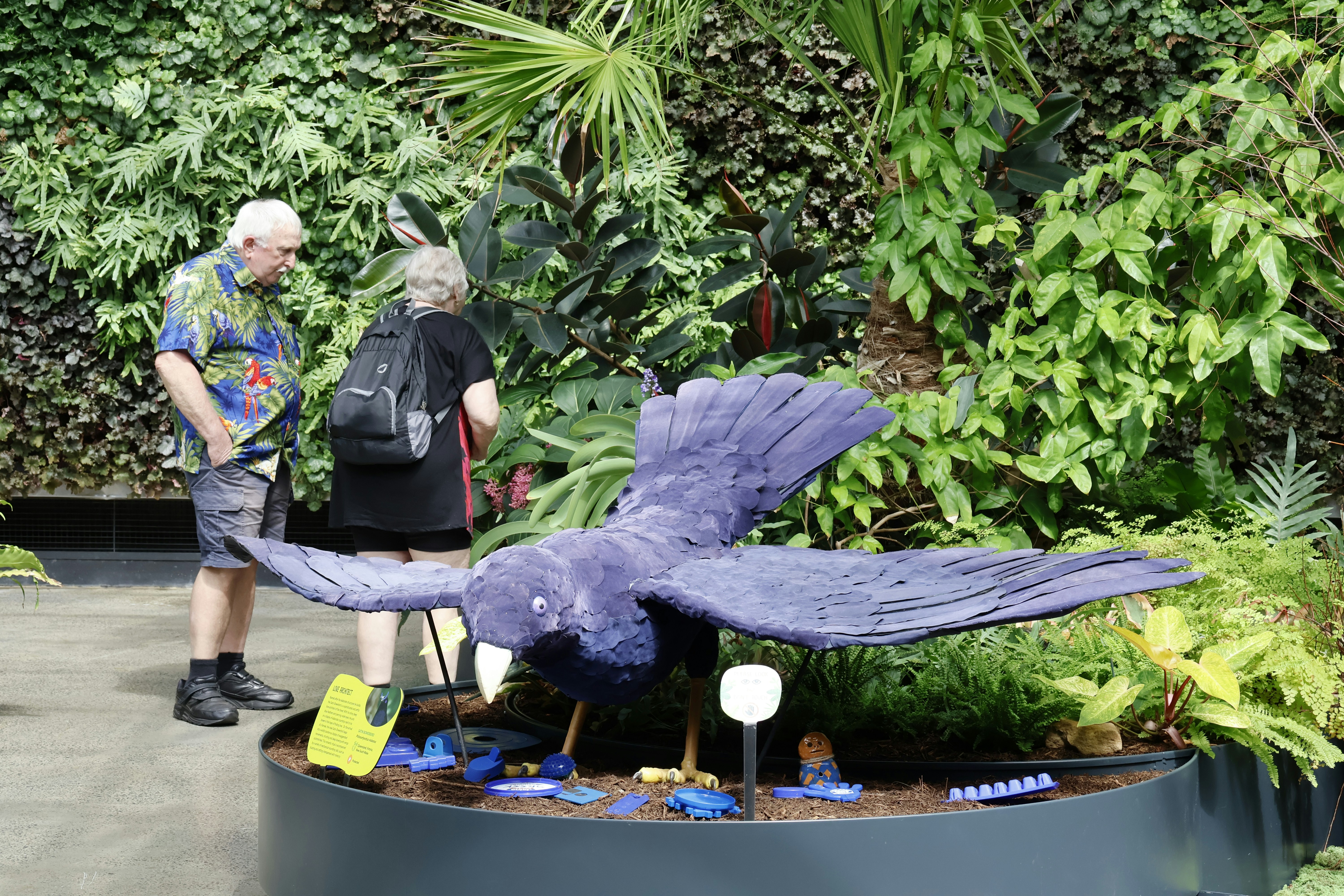Robust Blue Bird Sculpture Framed by Visitors and Lush Foliage