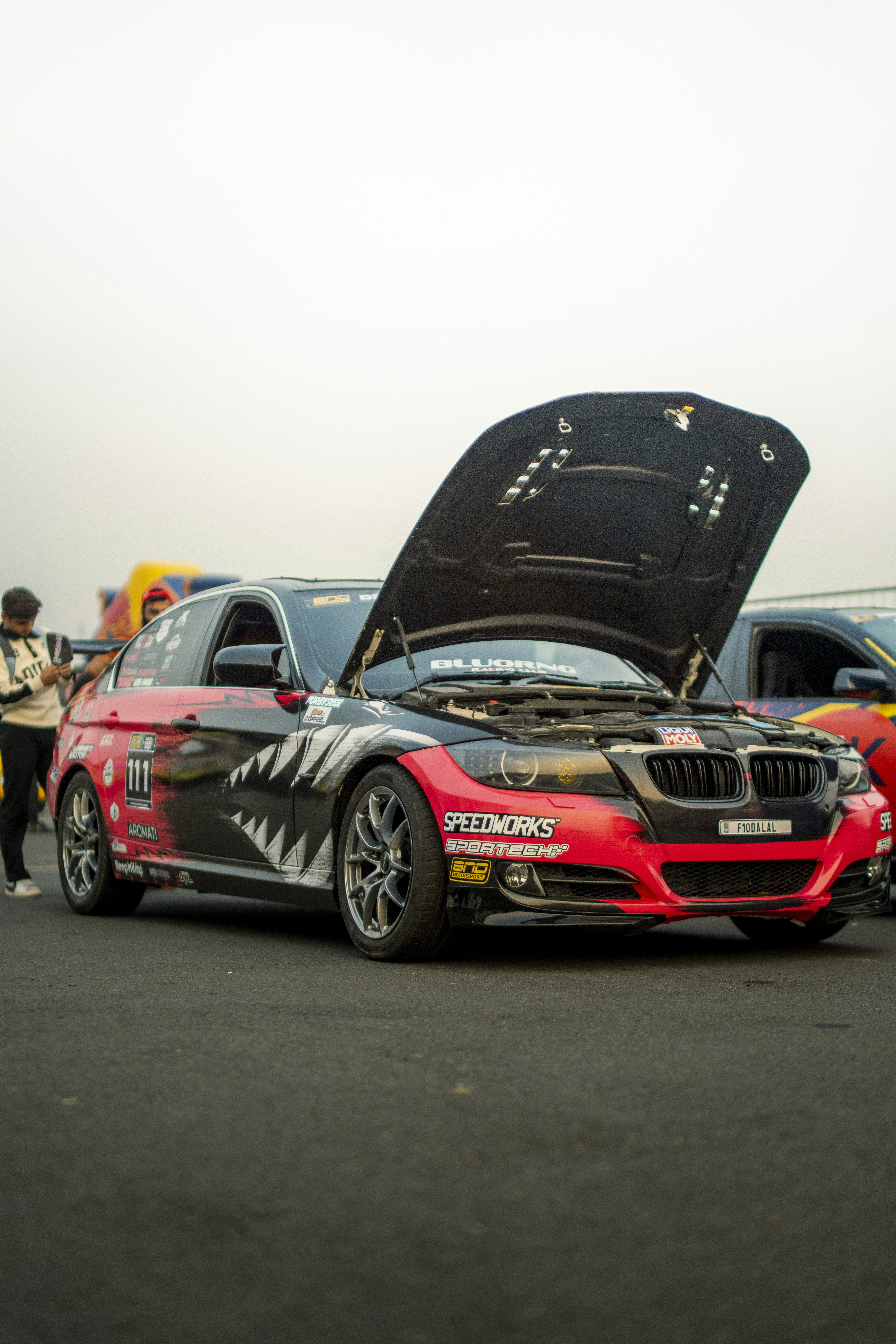 A red-and-black drift car with its hood open sits on a paved surface at a car show, others and a tent visible in the background. The scene focuses on the vehicle's front end and detailed decals.