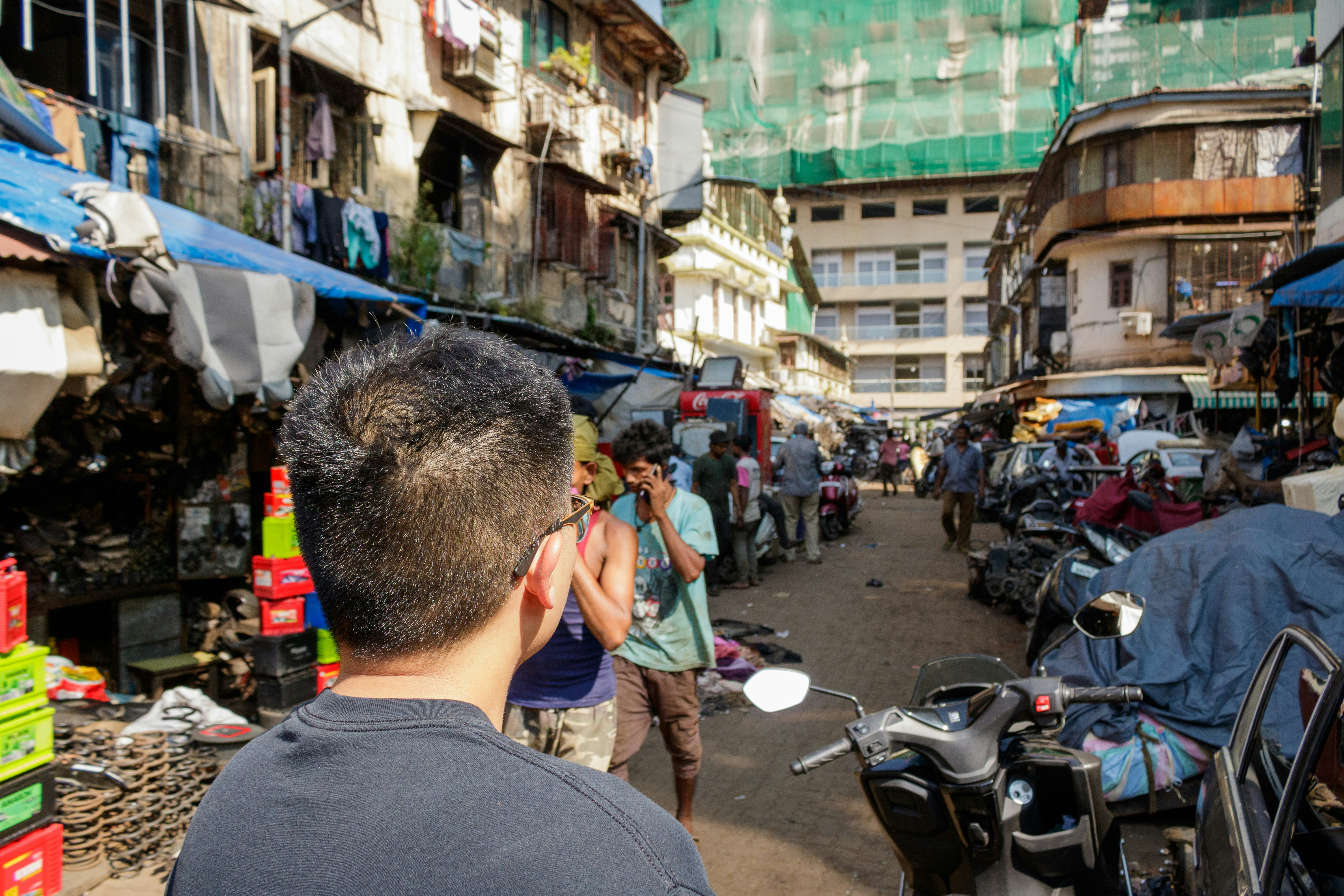 A visitor's perspective of a narrow lane in Mumbai's Chor Bazaar. In the foreground, shops under blue tarps display stacked red and green battery boxes and metallic springs. The street is occupied by pedestrians and parked scooters. Dilapidated residential structures with hanging laundry face a modern building under construction covered in green safety netting. This image is ideal for editorial use covering urban development, Indian commerce, or travel.