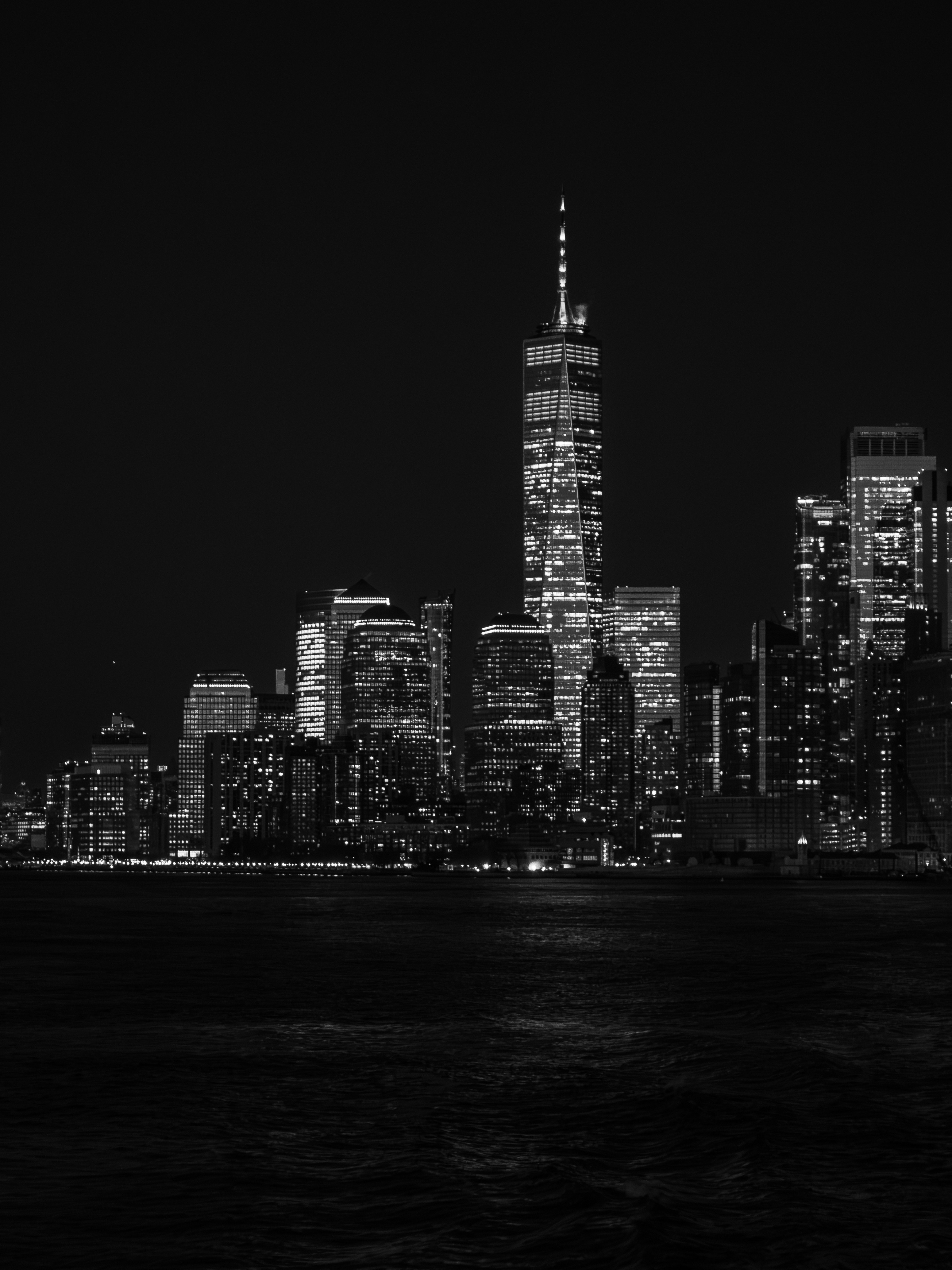 The Lower Manhattan skyline at night. It prominently features One World Trade Center, the tallest building in the United States at 1,776 feet, which officially opened in 2014. This factual cityscape is suitable for editorial use in articles regarding New York City’s urban history, architecture, or as large-format monochrome wall art.