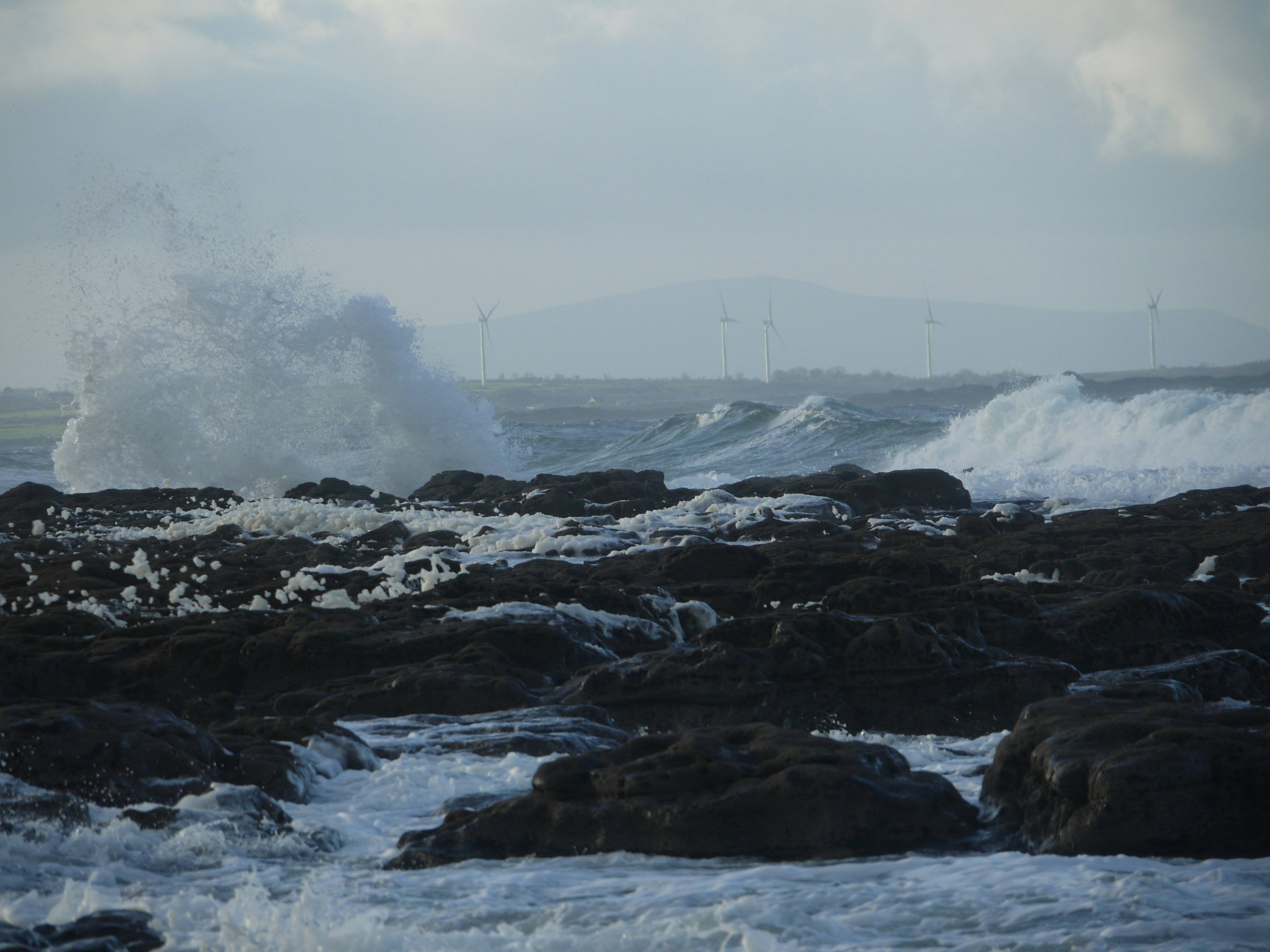 Rugged Coastline: Breaking Waves Against Dark Lava Flats Under Distant Wind Turbines