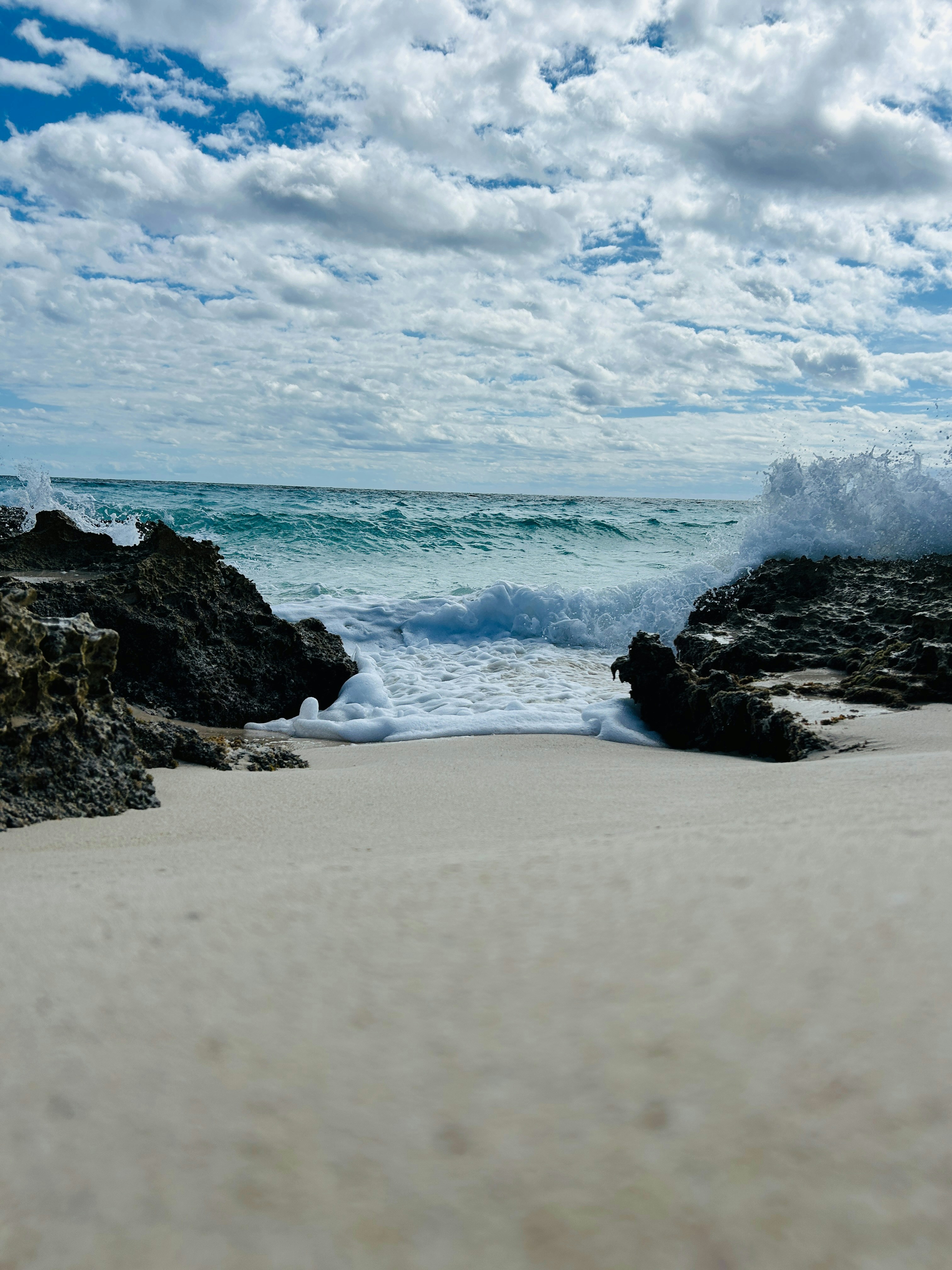 Snow-white foam wraps against rugged rocks at a sunlit beach leading to turquoise sea beyond.