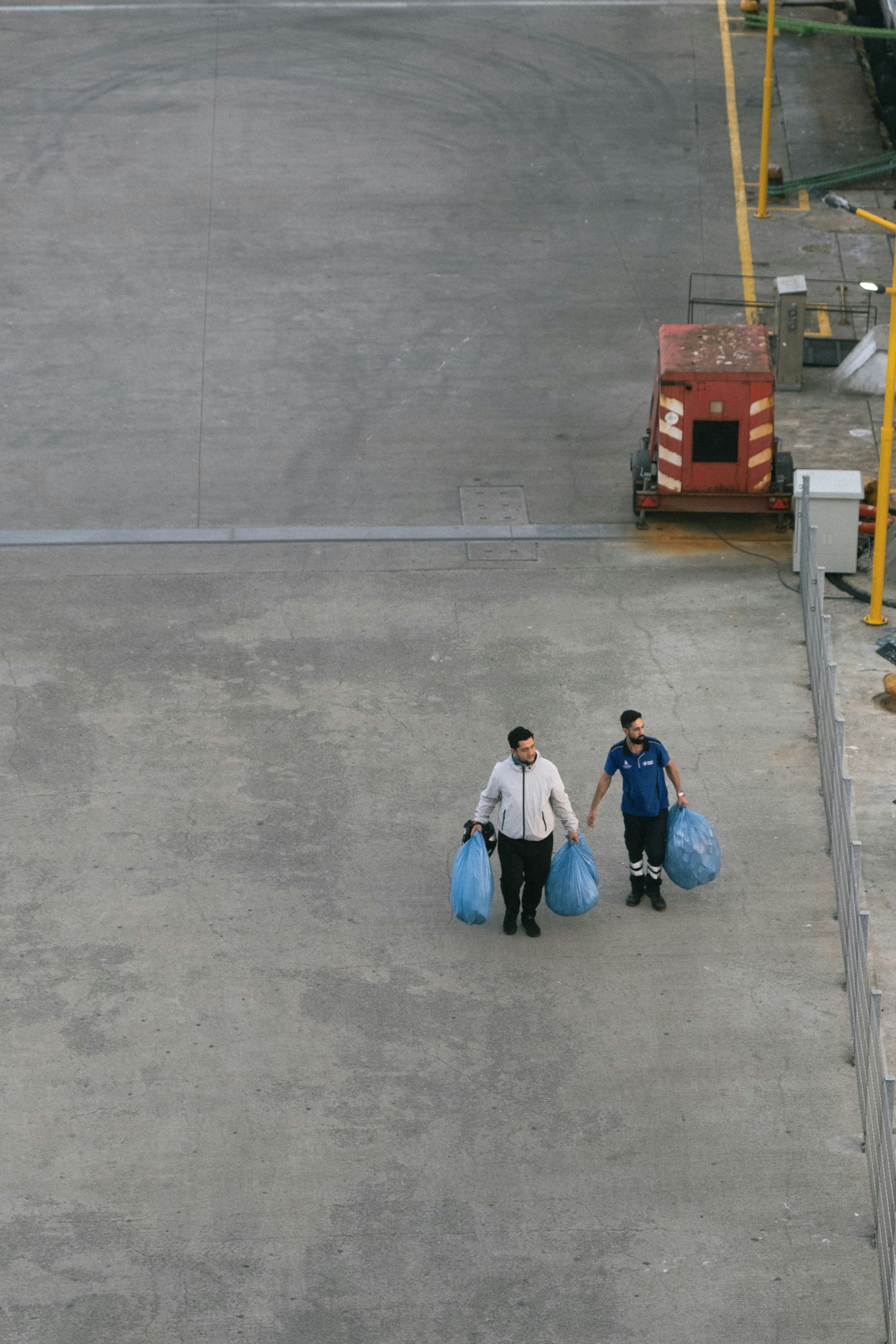 Two waste-collectors stroll across an empty industrial yard with blue bags in hand