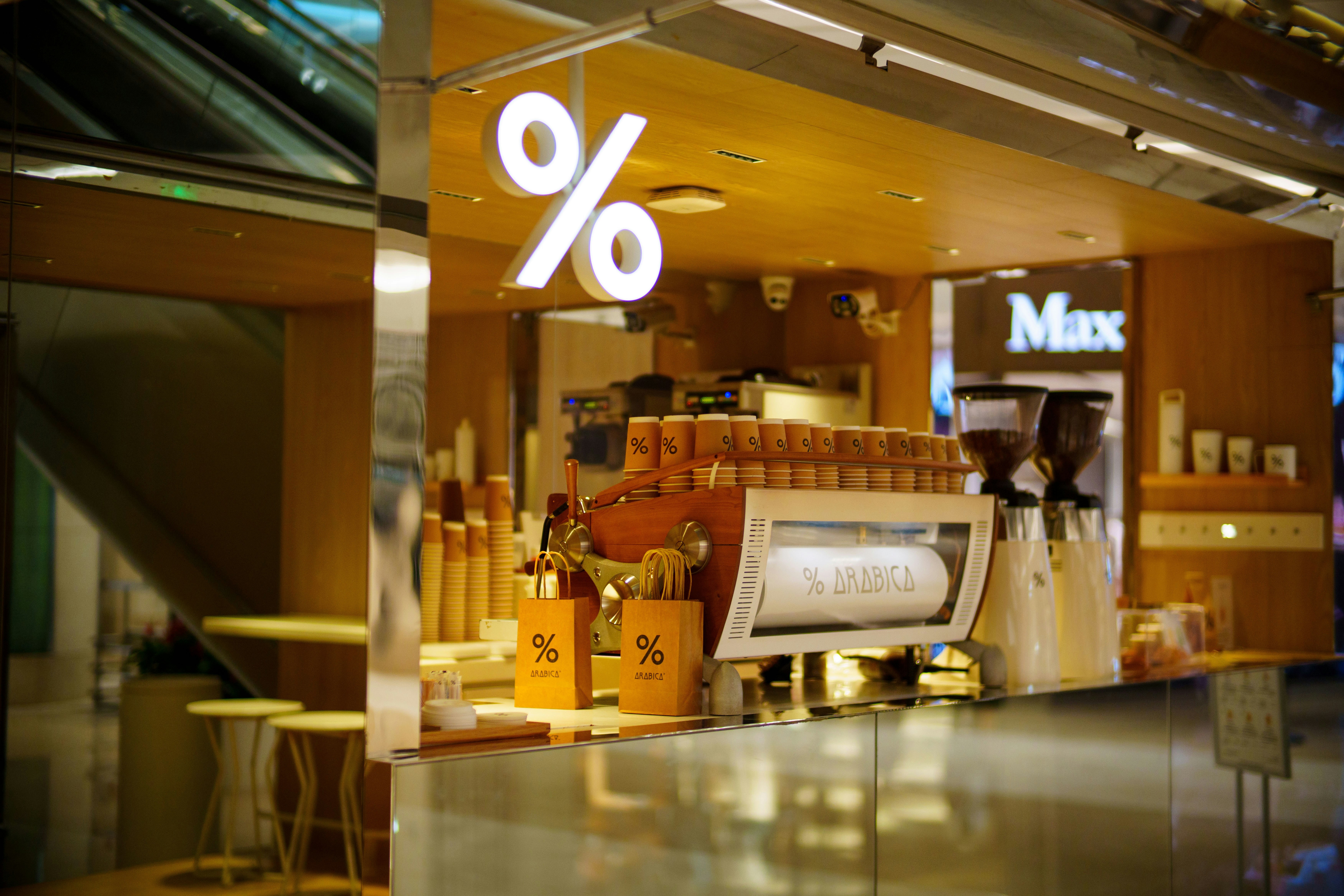 Bright café counter with a large percent symbol overhead; coffee equipment and take-away bags visible, suggesting a promotional display or retail coffee bar inside a shopping area.