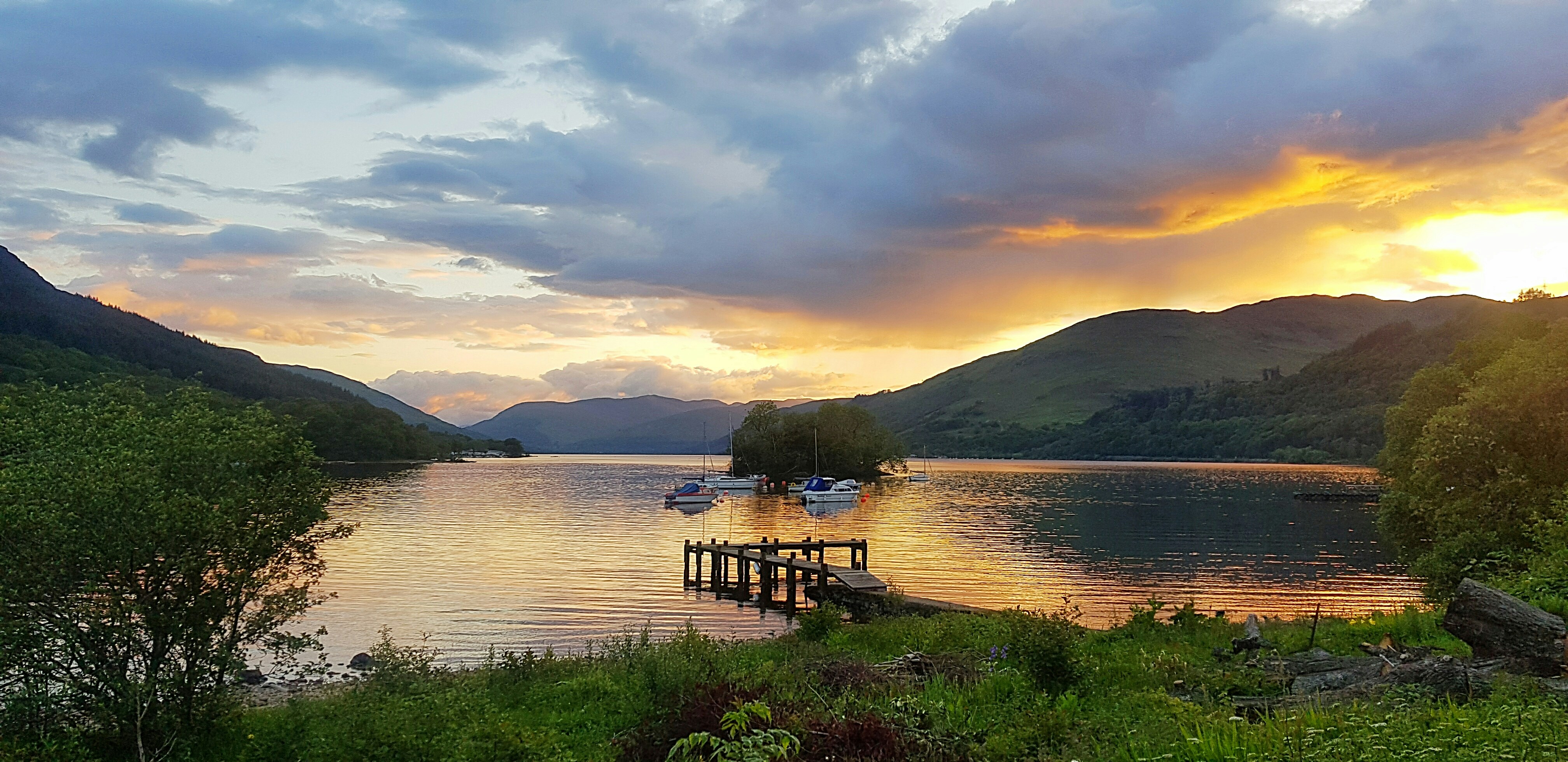 Sunset over a tranquil lake with a small dock and boats, framed by rolling hills and trees; a calm, landscape scene with warm light.