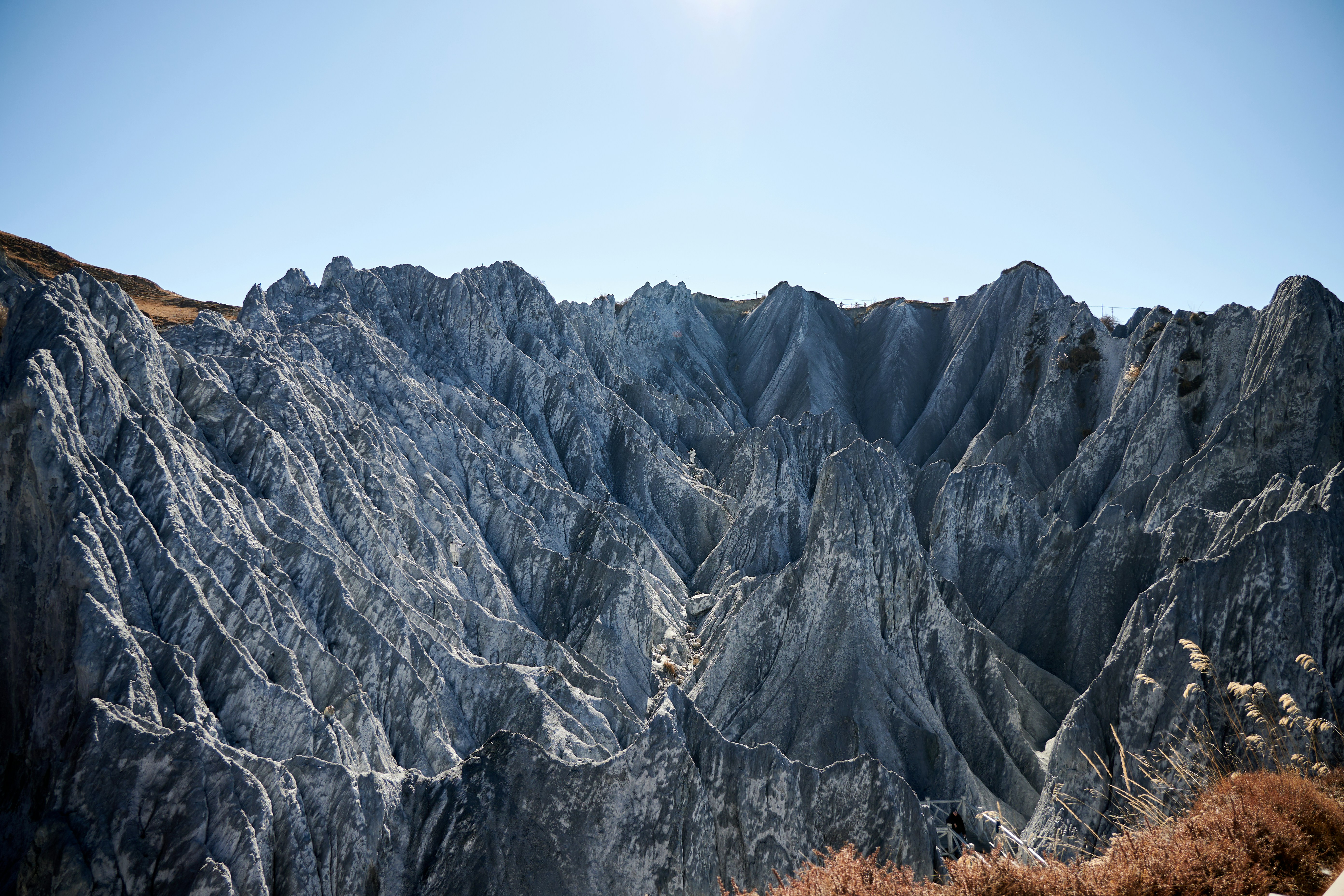 Jagged, eroded ridges of pale rock form a dramatic valley under a bright blue sky, with a touch of autumnal brush at the slopes' edge.