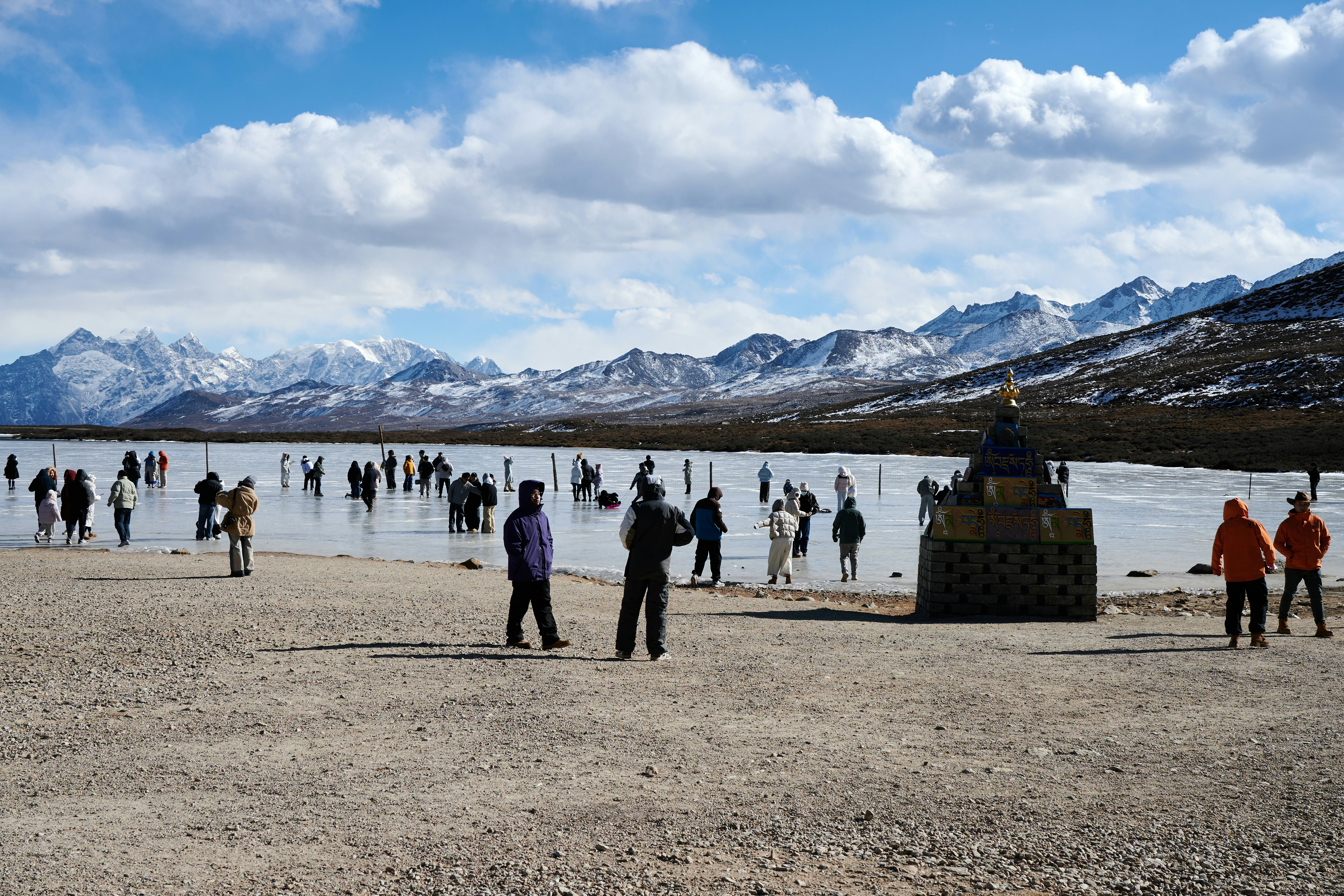 People on a snowy lakeshore with distant mountains and a stacked stone sculpture