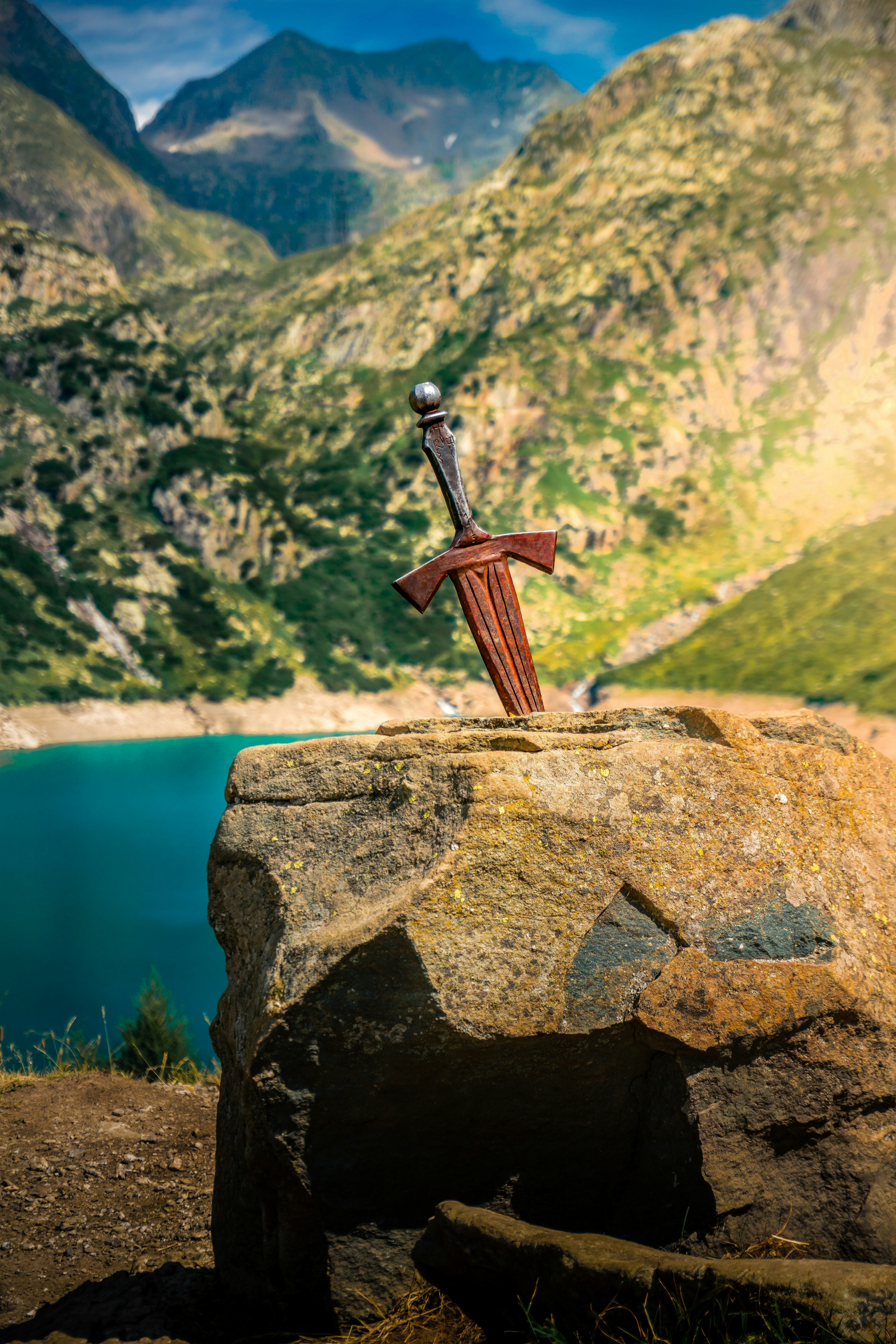 Rusty dagger planted in a stone pedestal overlooking a turquoise alpine lake and rugged mountains. The composition centers the relic with a blurred natural background and a sense of stillness.