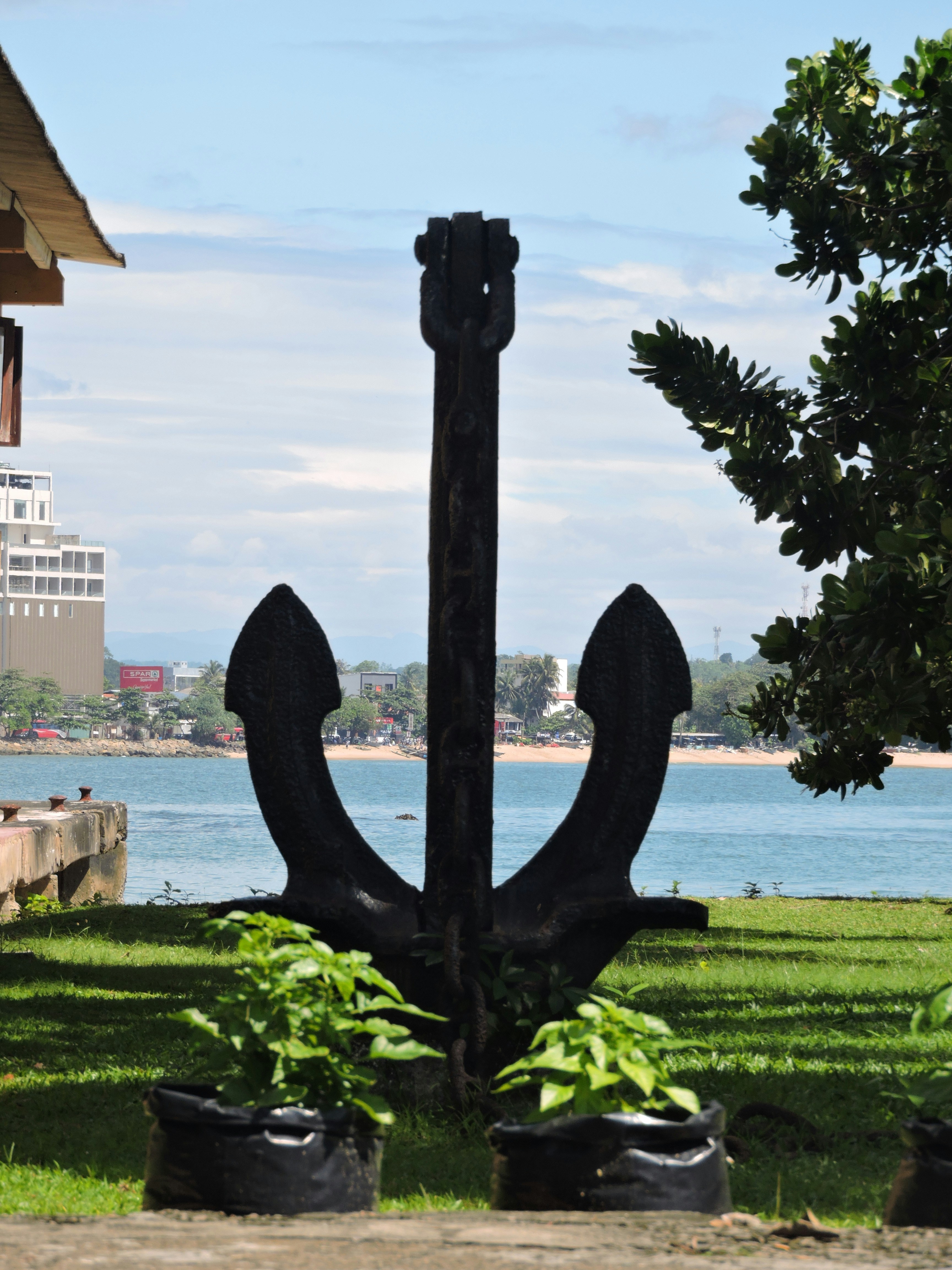 Large black anchor on grassy shore with buildings.