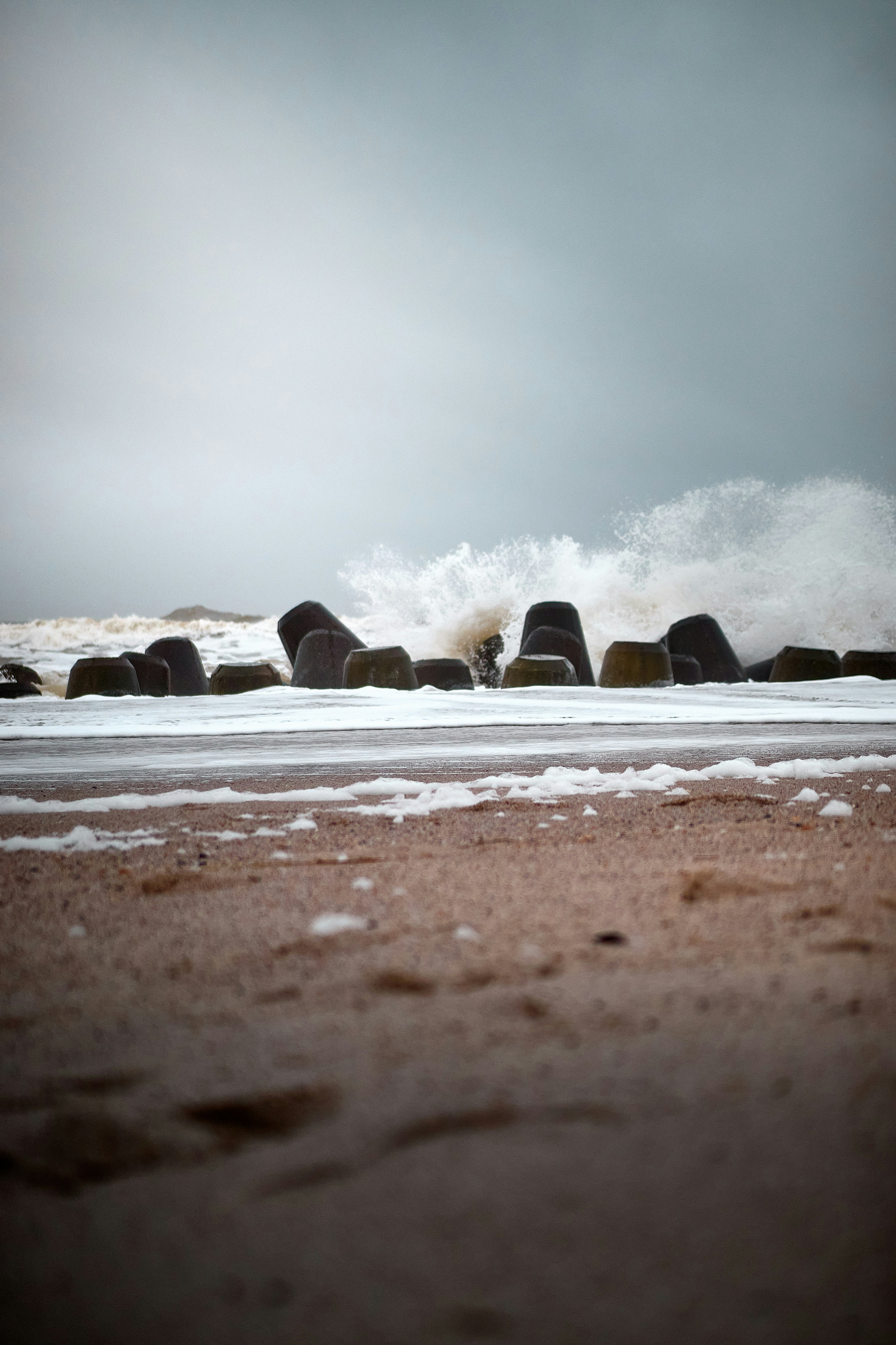 Waves crash against rocky breakwaters along a sandy beach, captured with a low-angle perspective showing frothy spray and wet foreground. The scene emphasizes the contrast between water and shore detail.